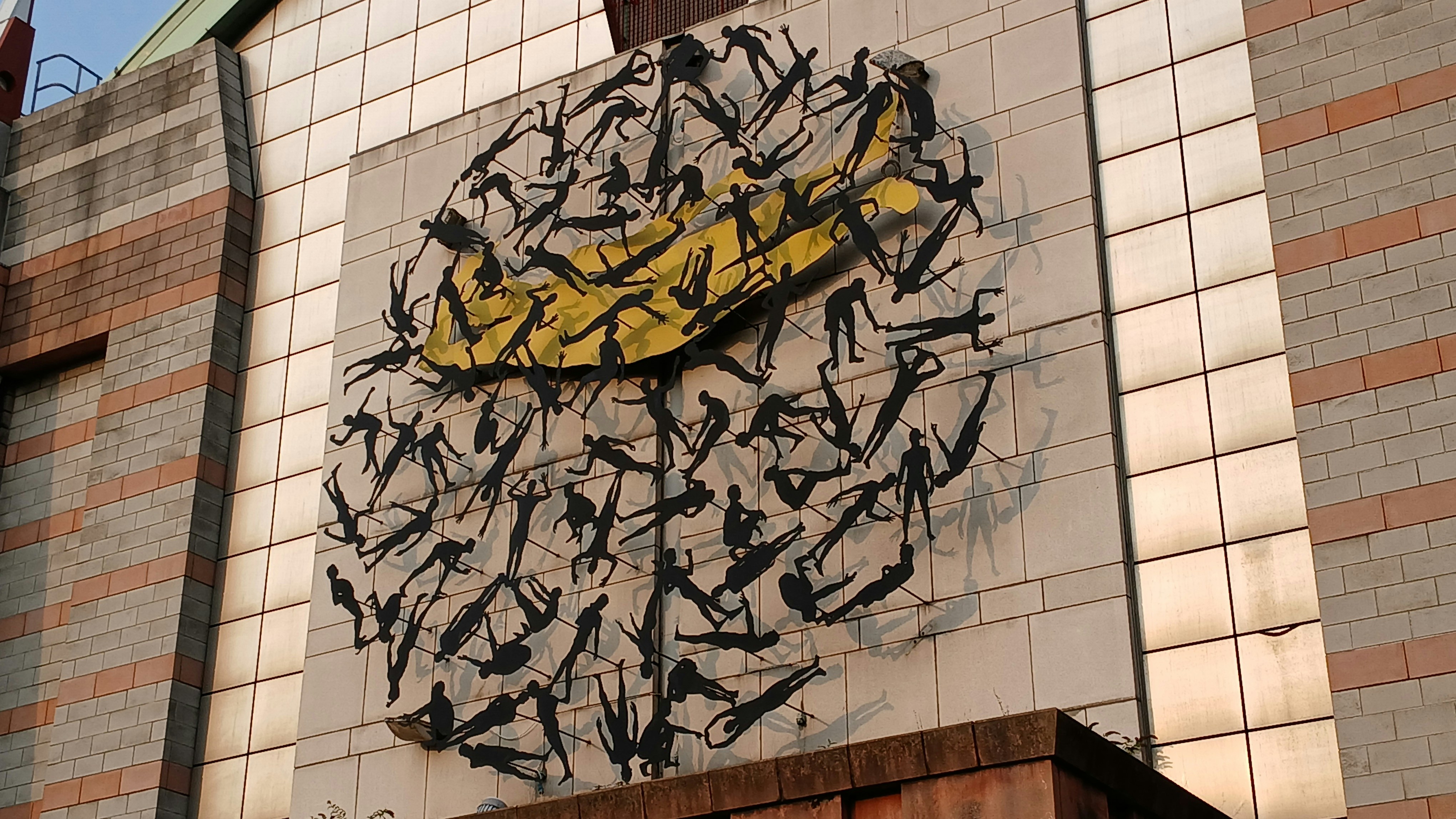 Circular mural installation of black silhouettes spiraling around a yellow core on a brick façade.