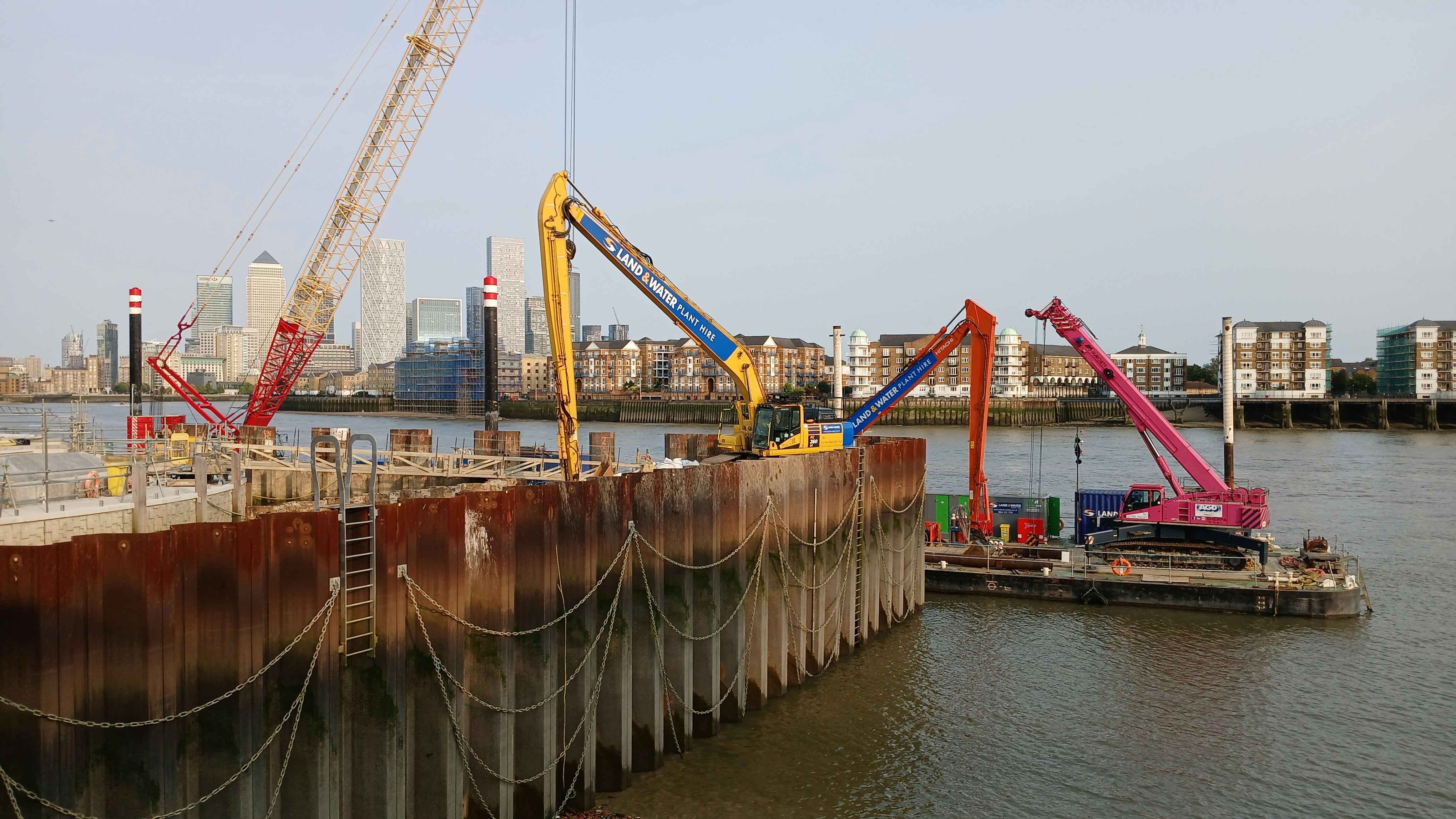 A rusted barge with colorful cranes moored along a river, with a distant city skyline.