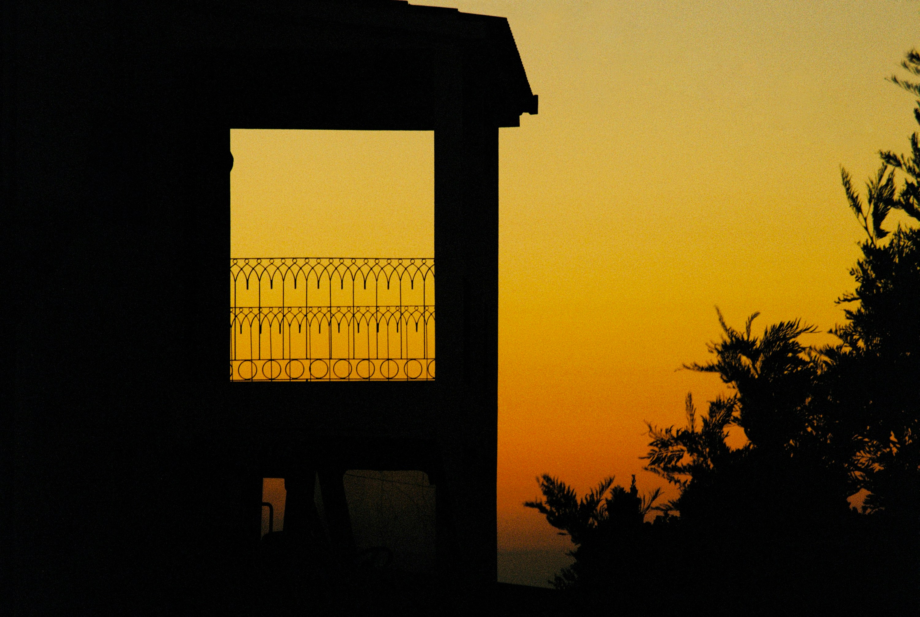 Silhouette of a two-story structure with a decorative balcony window set against a gradient orange sunset sky. The warm glow within the window contrasts with the dark facade, emphasizing architectural detail.