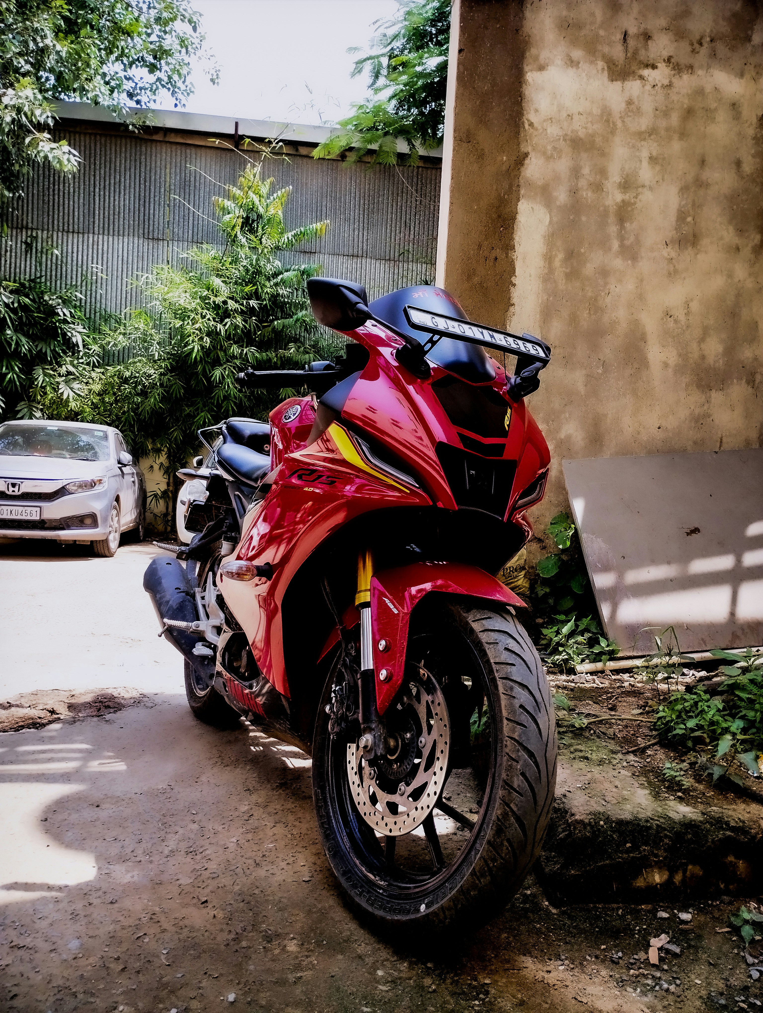 A red motorcycle parked in a sunlit courtyard, with a weathered wall and a car in the background. The bike's glossy finish and bold lines dominate the composition.
