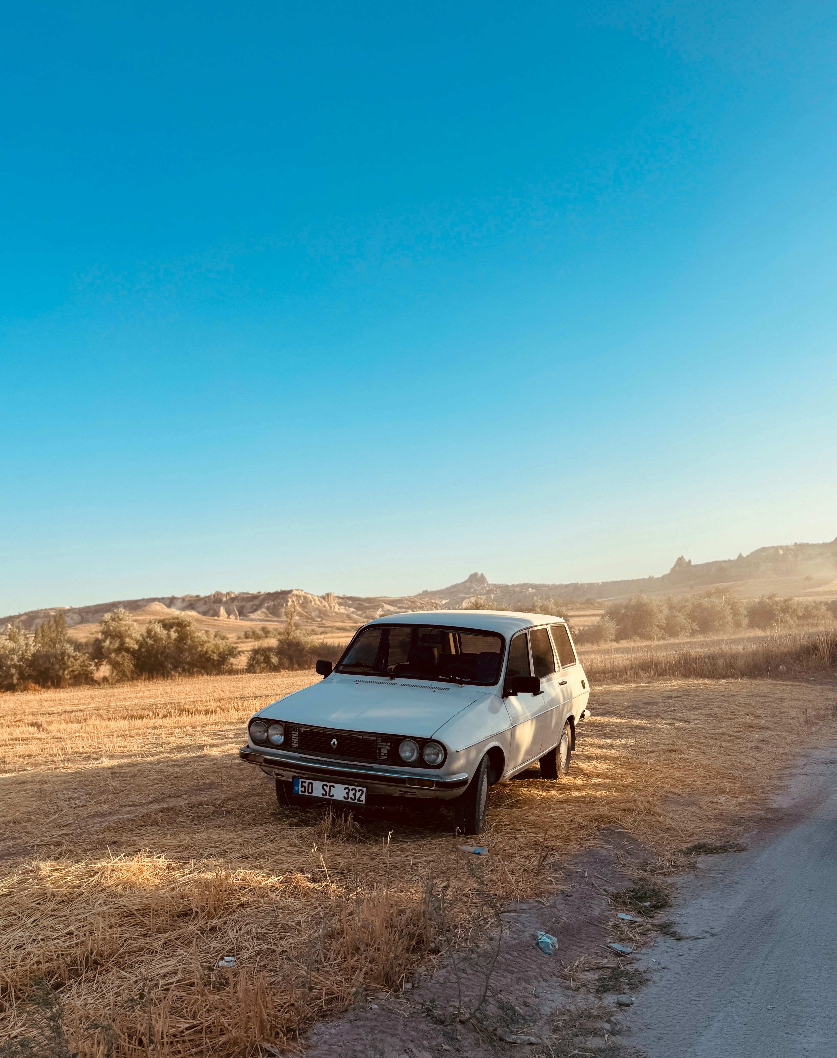 A vintage white car parked on a golden field under a clear blue sky, surrounded by rolling hills. The scene evokes nostalgia and a sense of adventure.