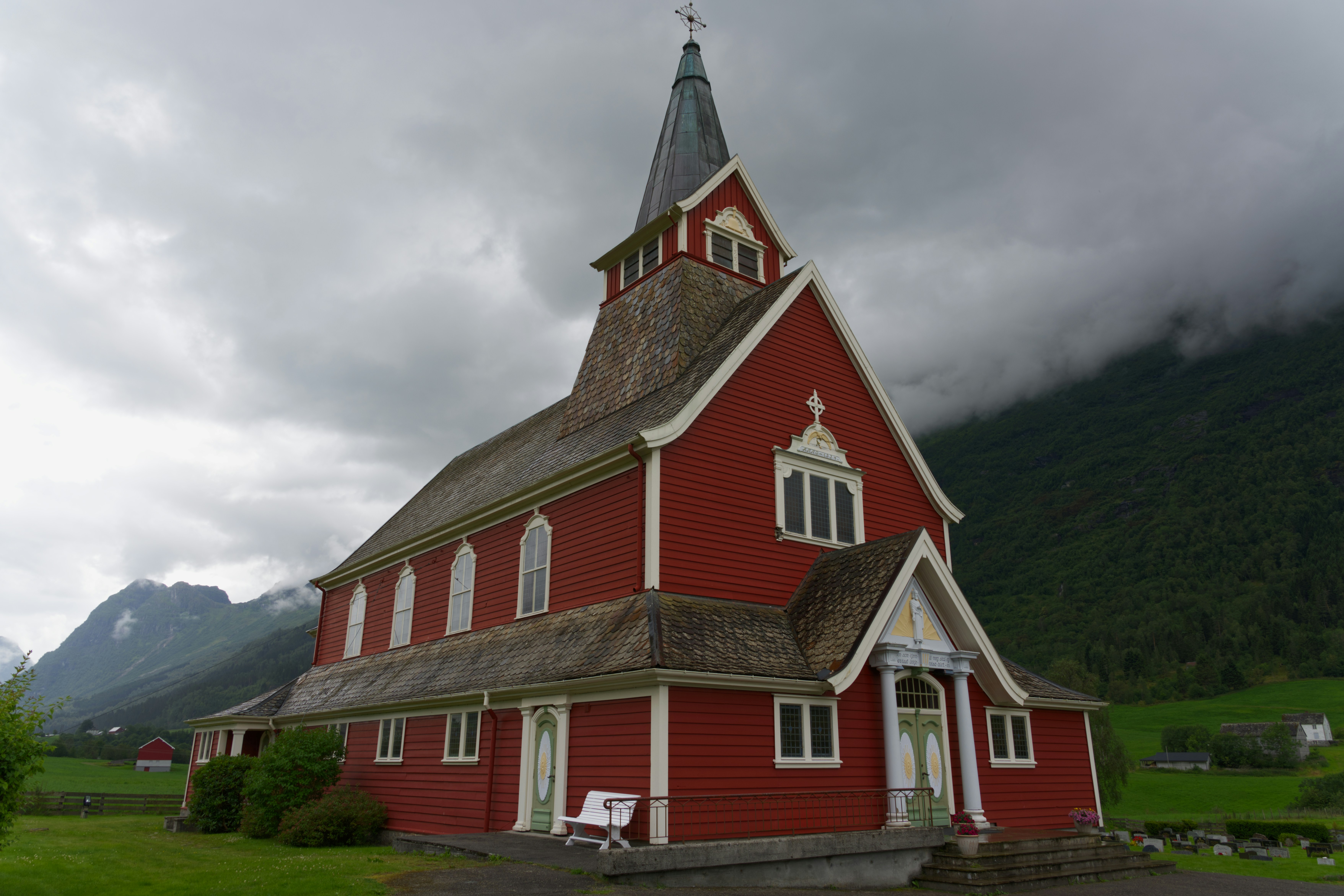 A striking red wooden church set against a dramatic mountainous backdrop, showcasing traditional Norwegian design elements. The scene captures the essence of rural heritage.