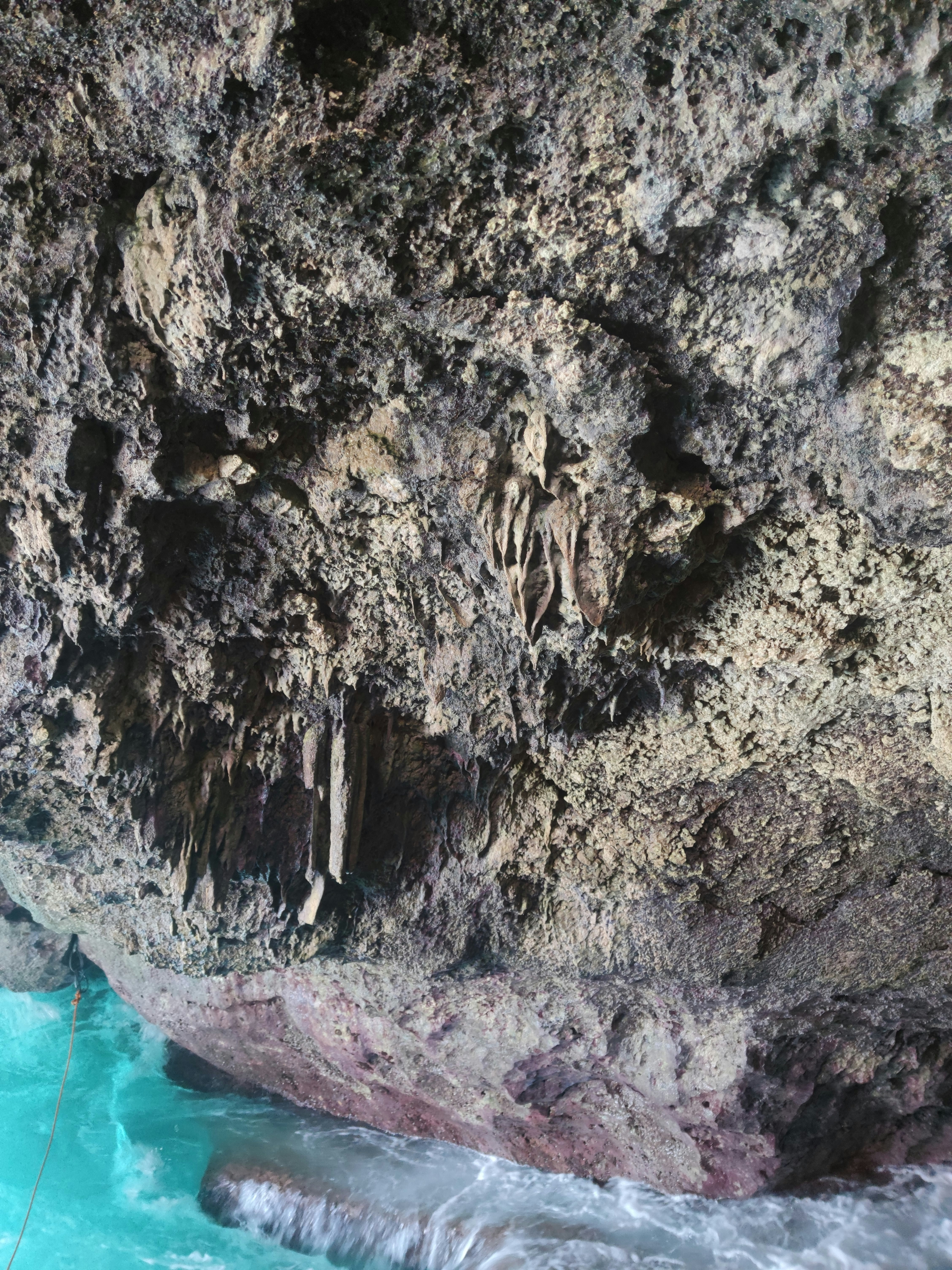 Rugged sea-cave wall with hanging stalactites above turquoise water along a rocky shoreline.