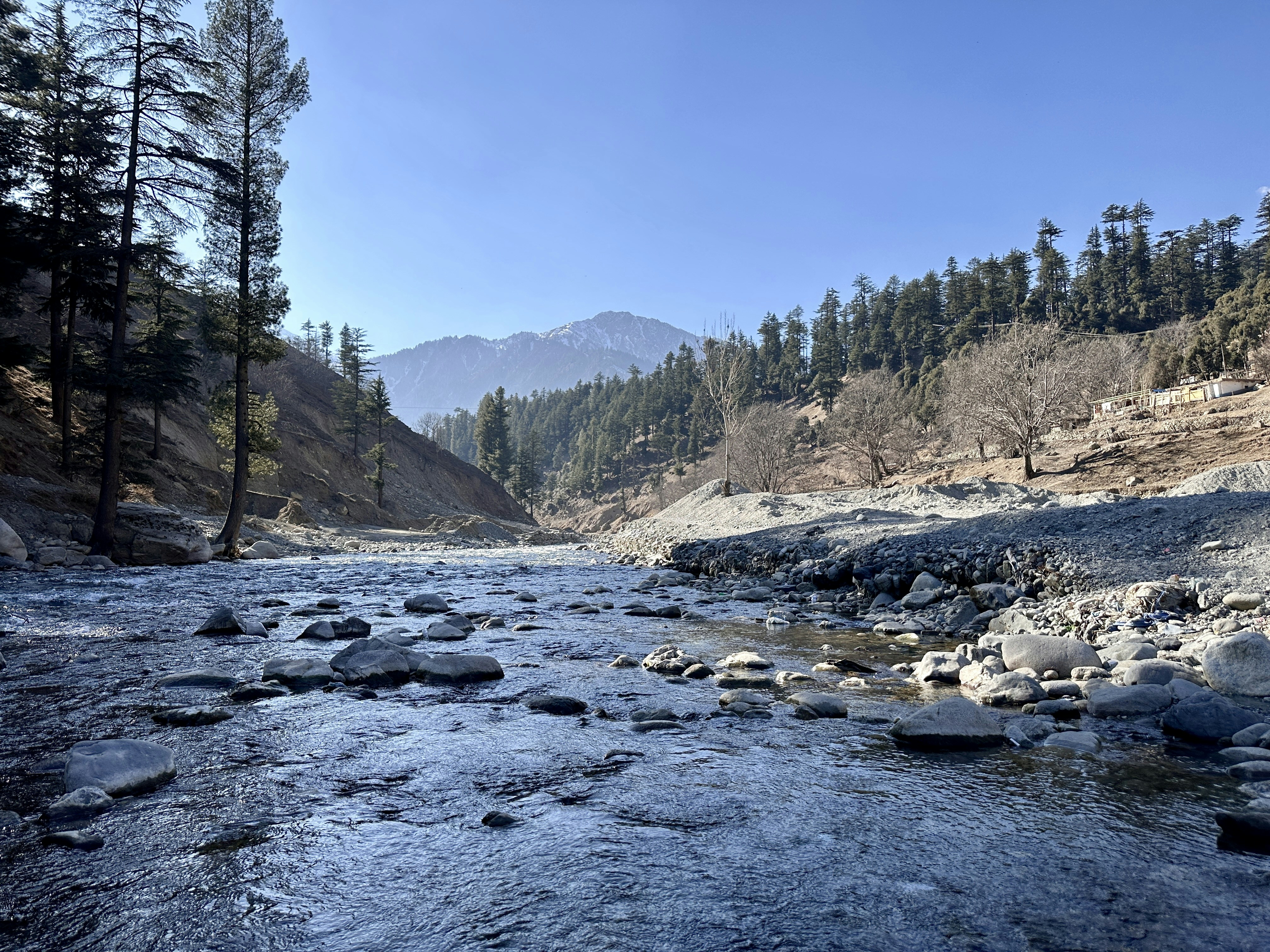 Clear river winding through a rocky landscape, flanked by coniferous trees and distant mountains under a bright blue sky.