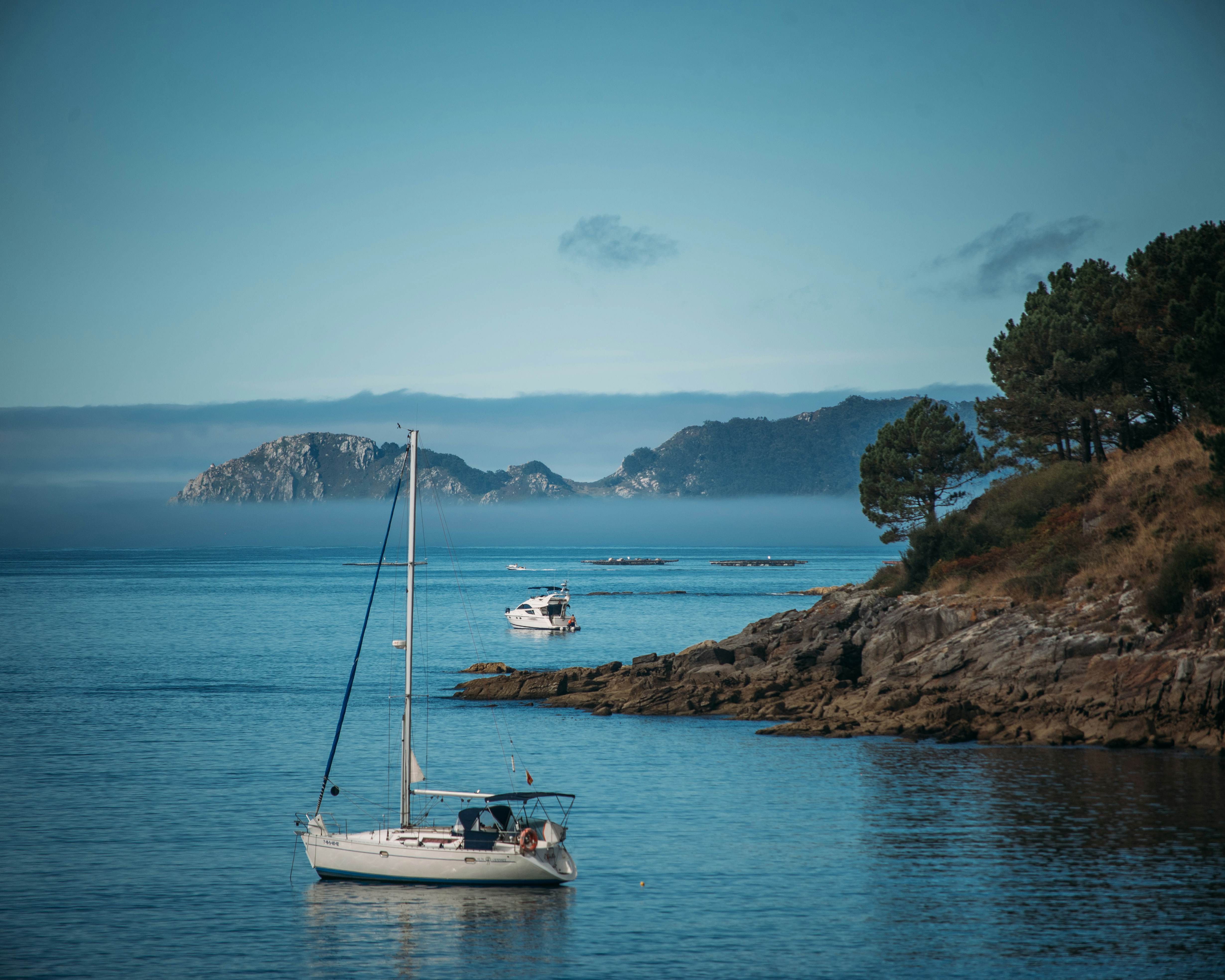 Sailboats drifting near rocky coastline under a clear blue sky.
