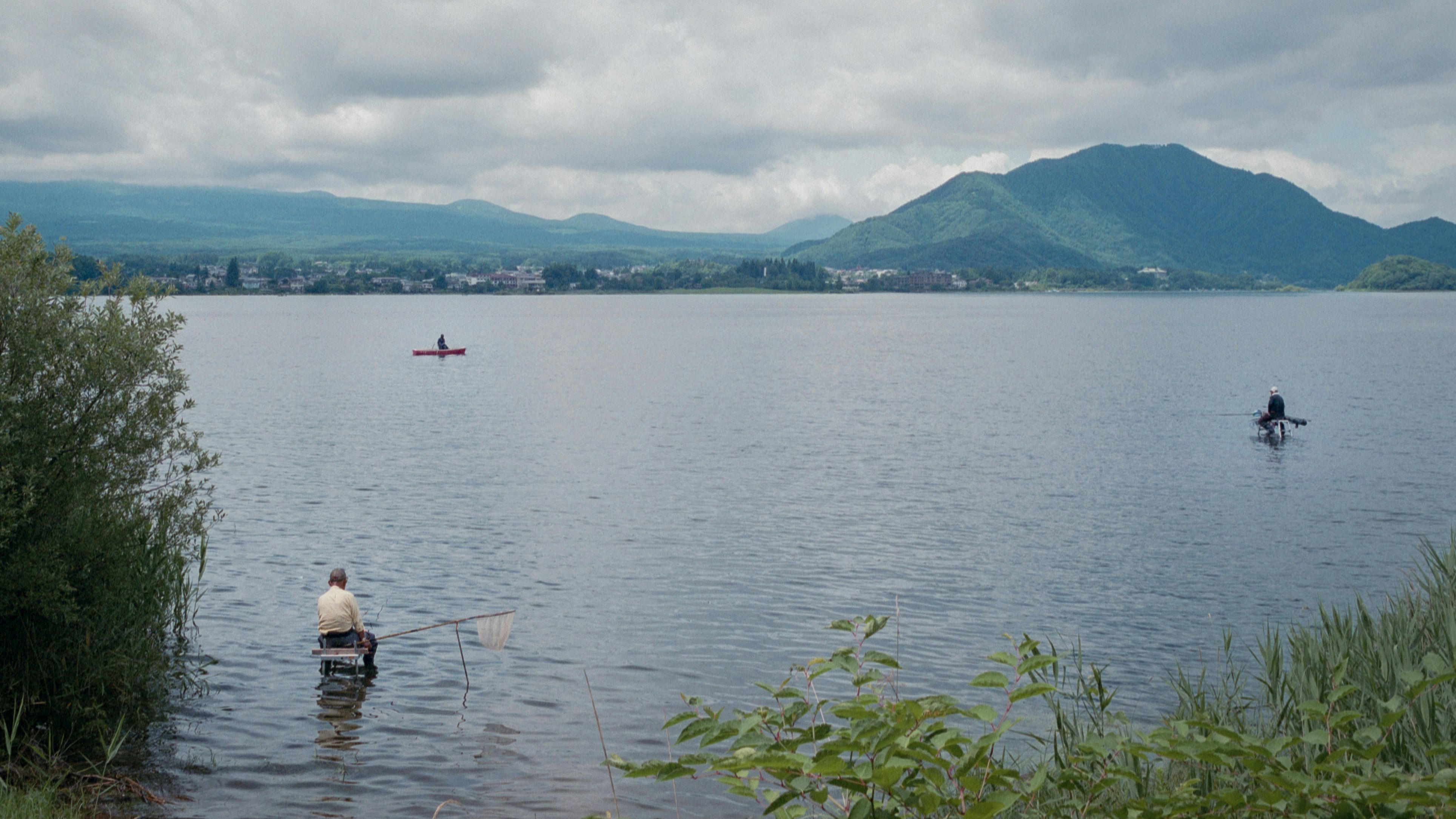 Person standing in a lake with a distant boat and mountains under a cloudy sky.