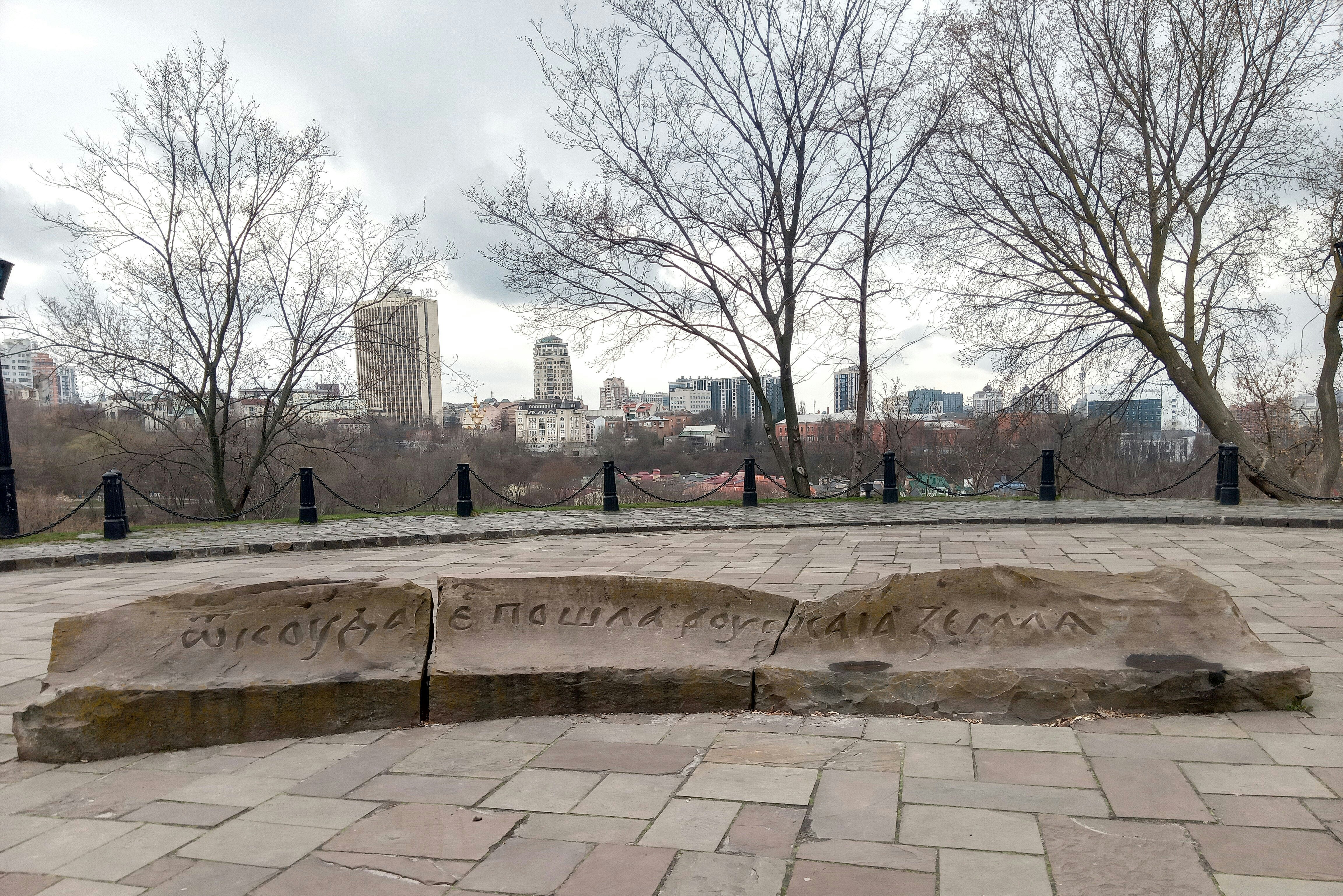 Engraved stone monument with inscriptions overlooking a cityscape, framed by bare trees. A cloudy sky adds a contemplative mood.