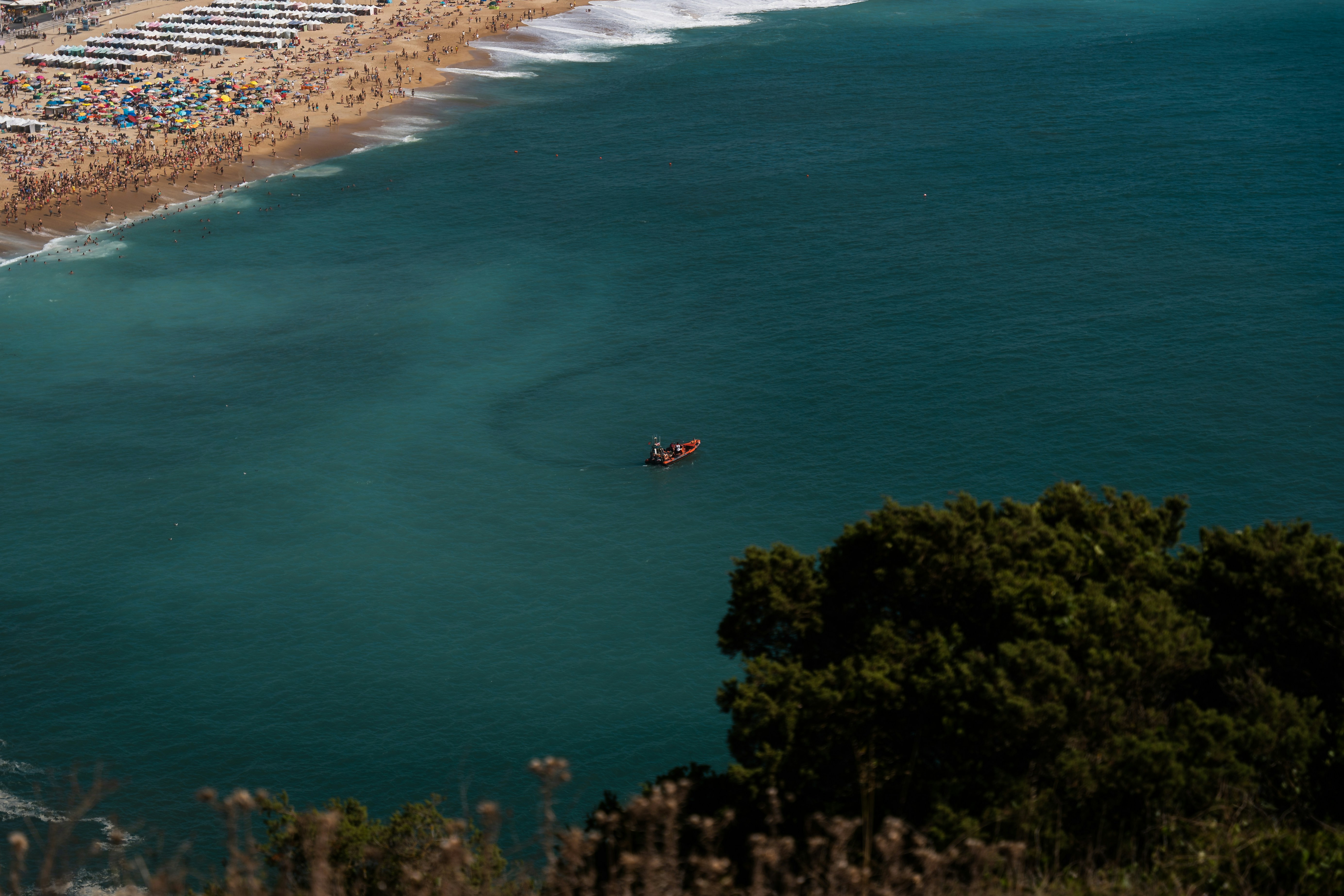 A lone fishing boat navigates the tranquil waters, framed by a bustling beach in the distance. The contrast between the calm sea and lively shore creates a striking visual narrative.