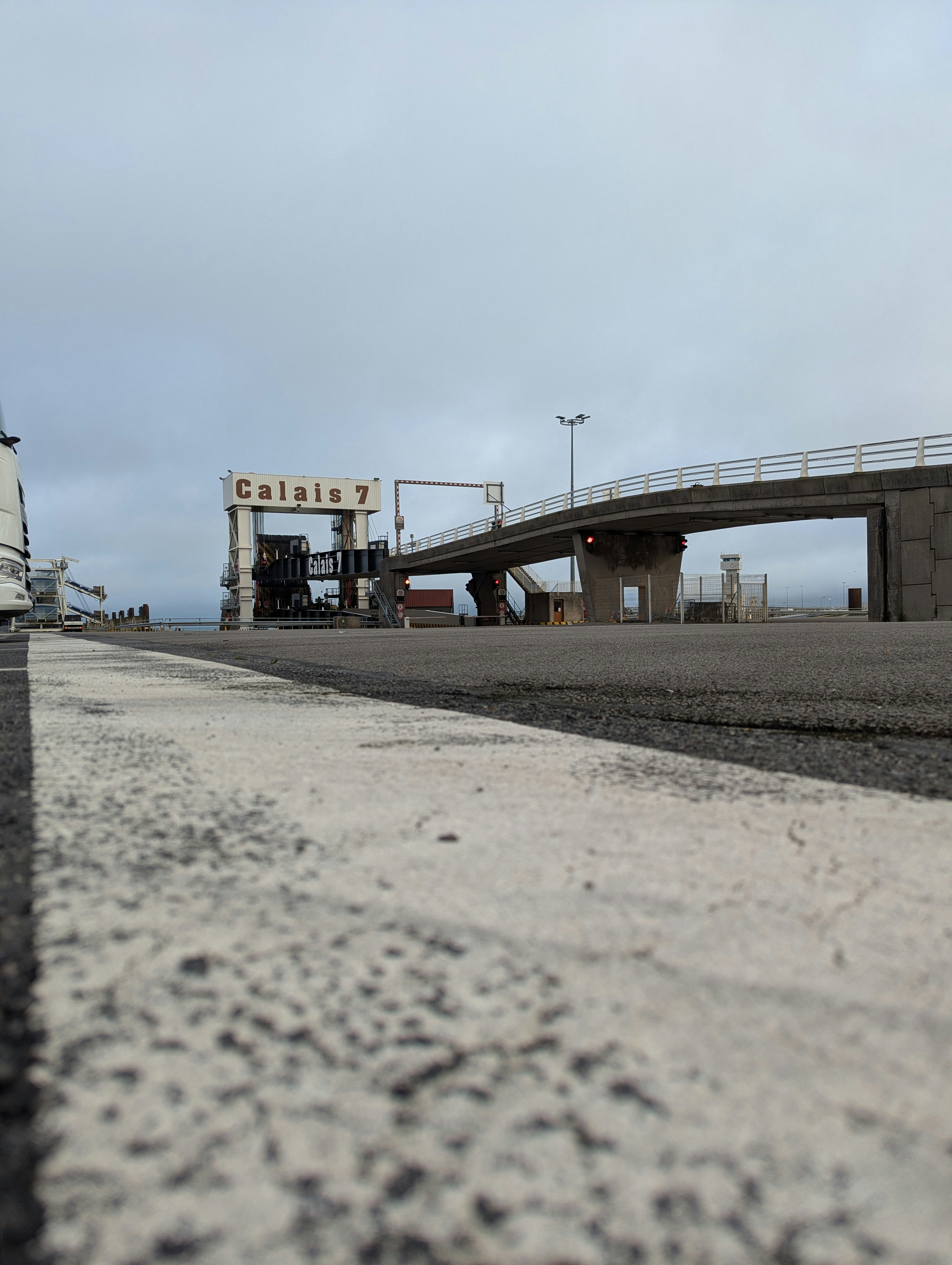 The Calais 7 ferry terminal entrance, showcasing the concrete structure against a cloudy sky, with a focus on the road leading to it.