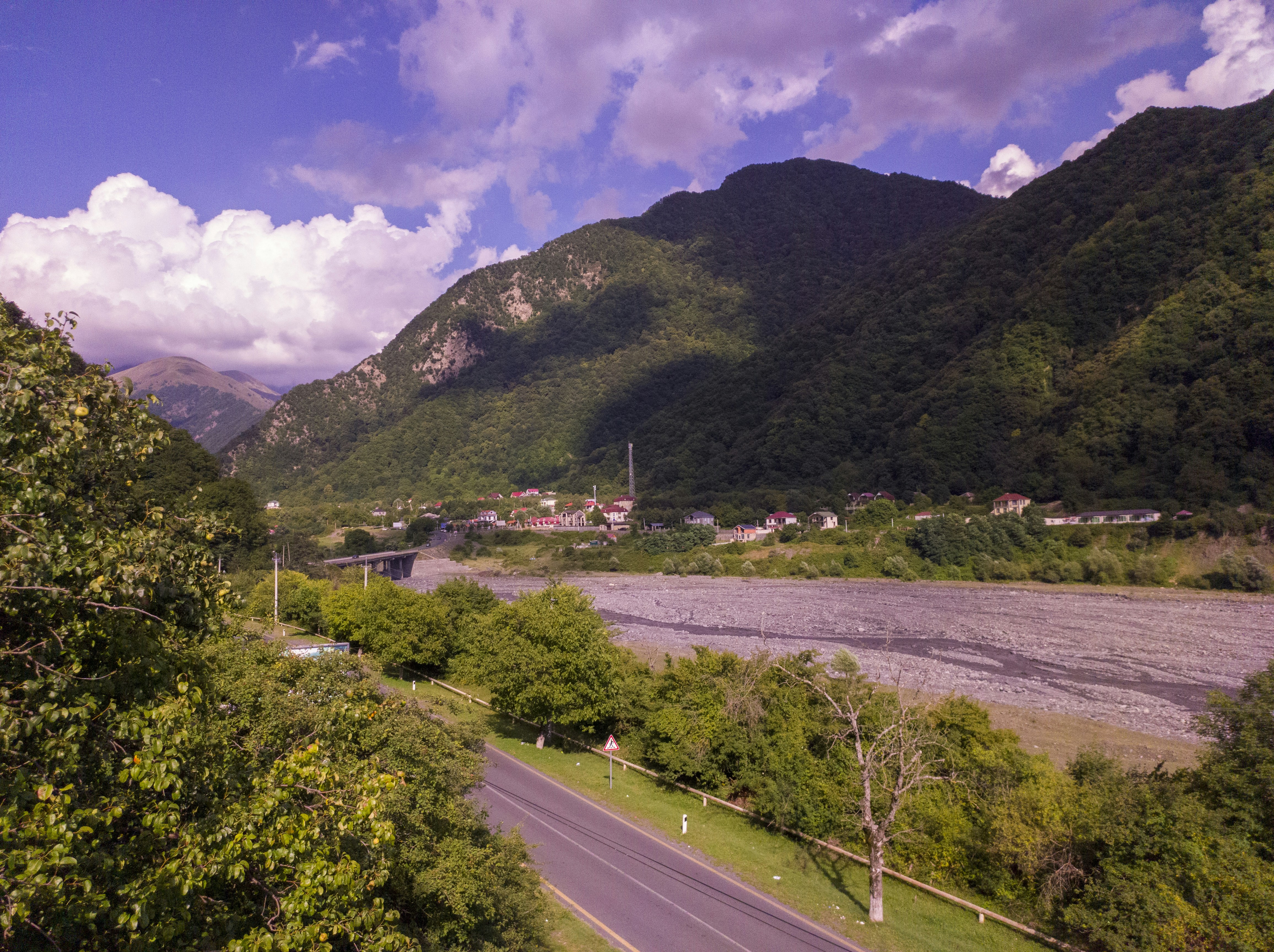 Landscape photograph capturing a river valley, roadside greenery, and a village at the base of forested mountains under a bright sky.
