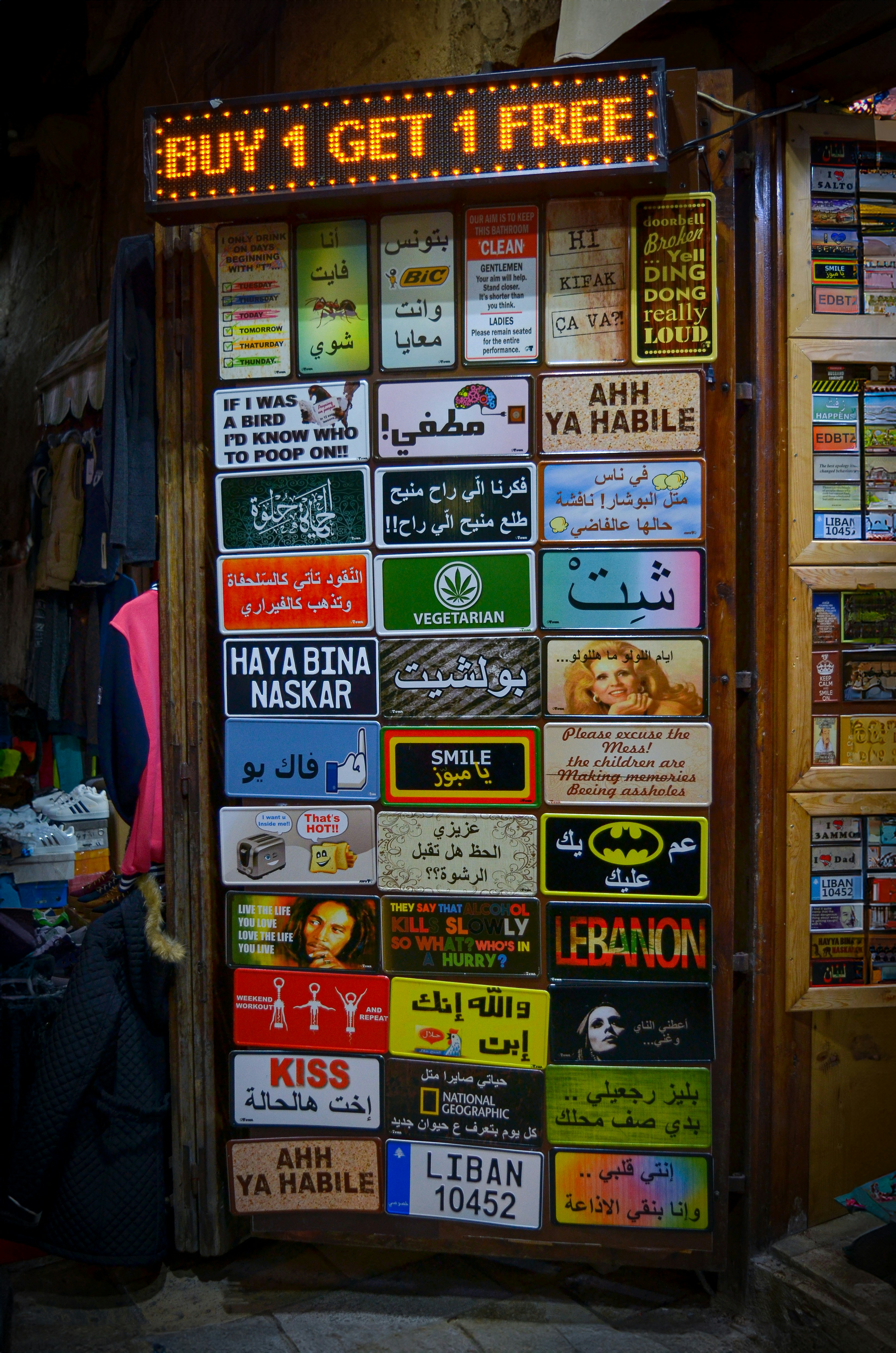 Colorful collection of humorous and informative signs displayed on a market stall, showcasing local culture and language. Bright neon 'BUY 1 GET 1 FREE' sign draws attention.