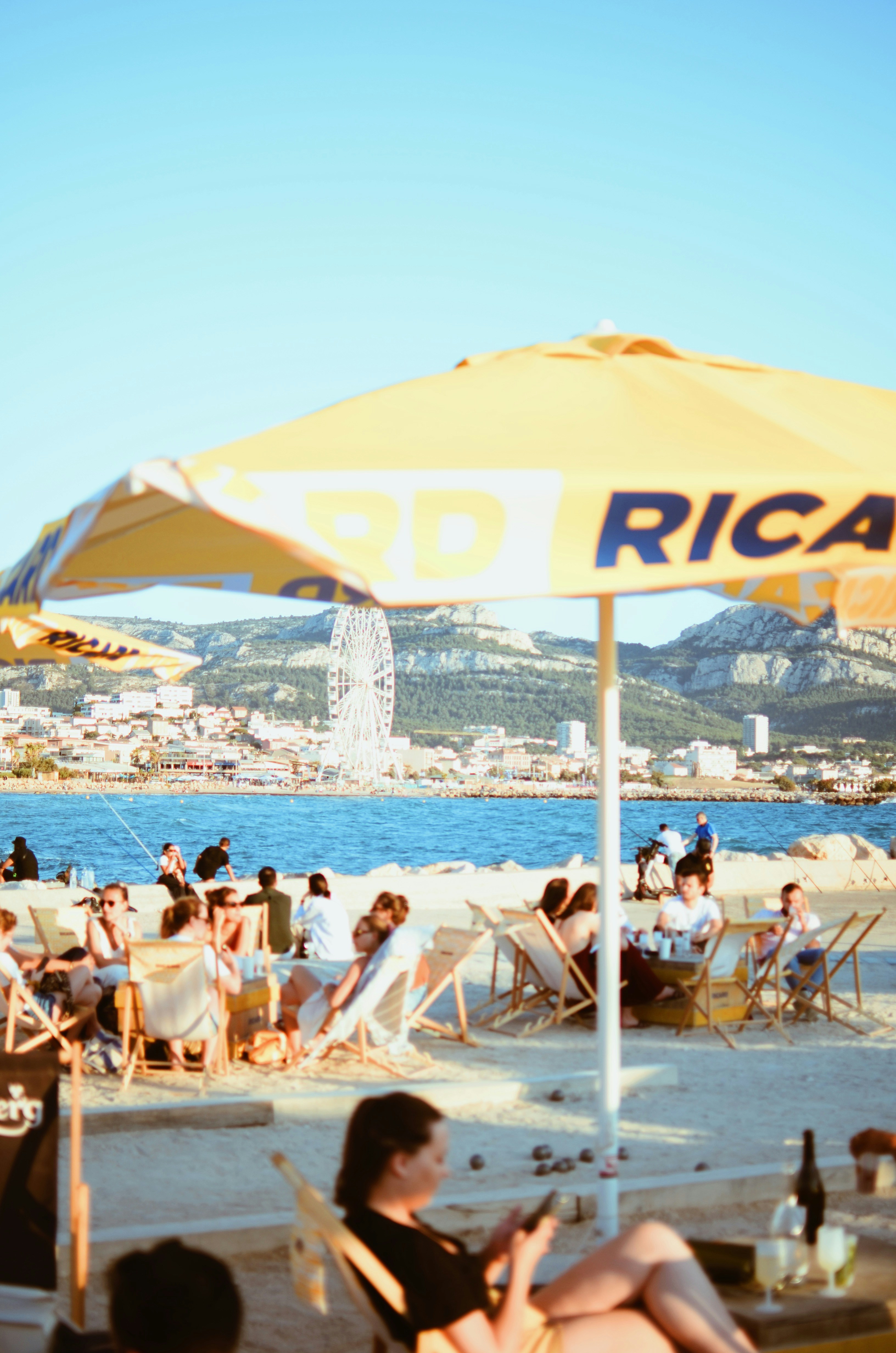 End of the day by the Mediterranean Sea in Marseille, with people relaxing on sun loungers and enjoying a drink. | A group of people sitting under umbrellas on a beach
