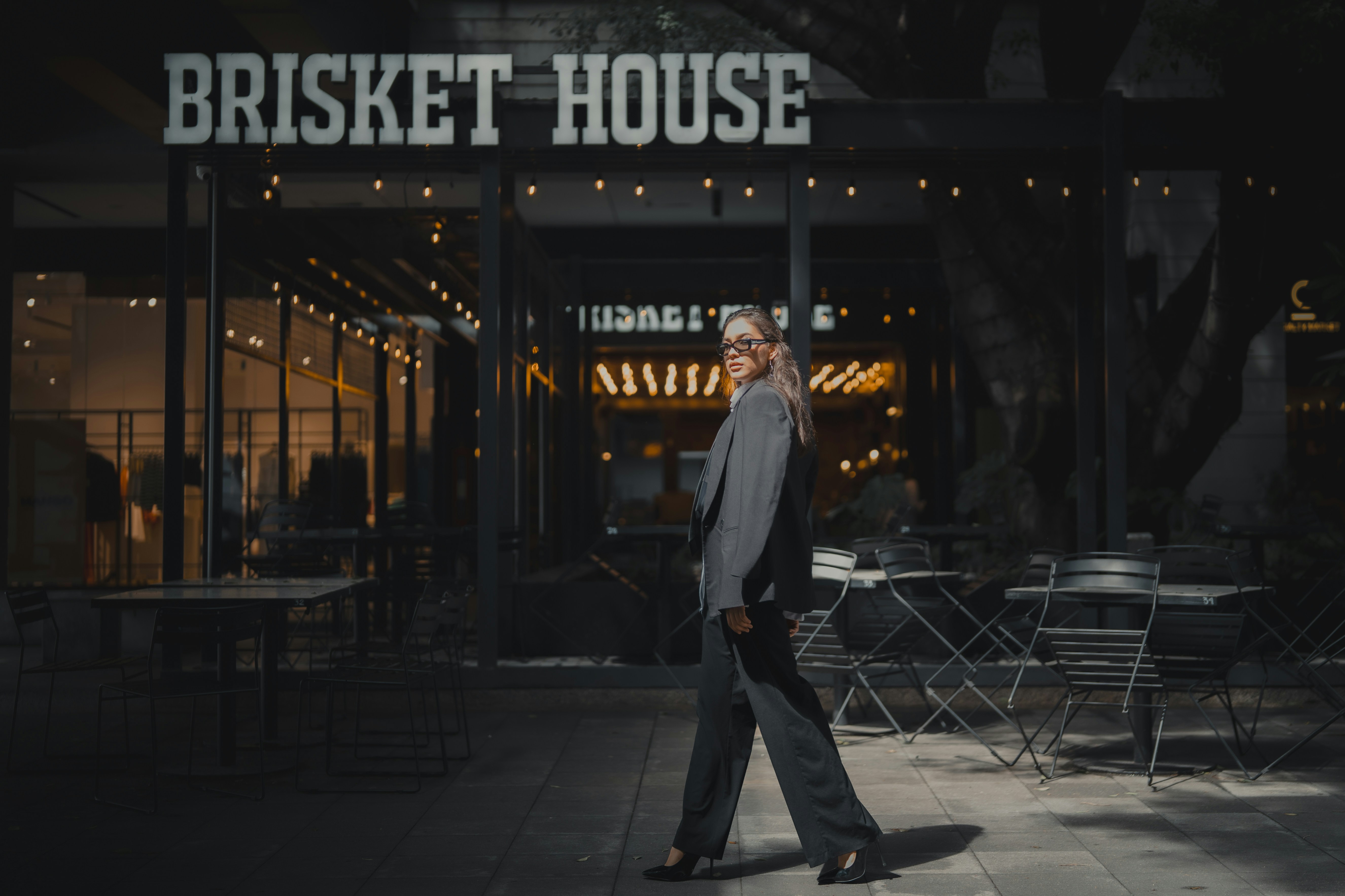 Person walking past a restaurant with 'Brisket House' sign, surrounded by outdoor seating and soft urban lighting.