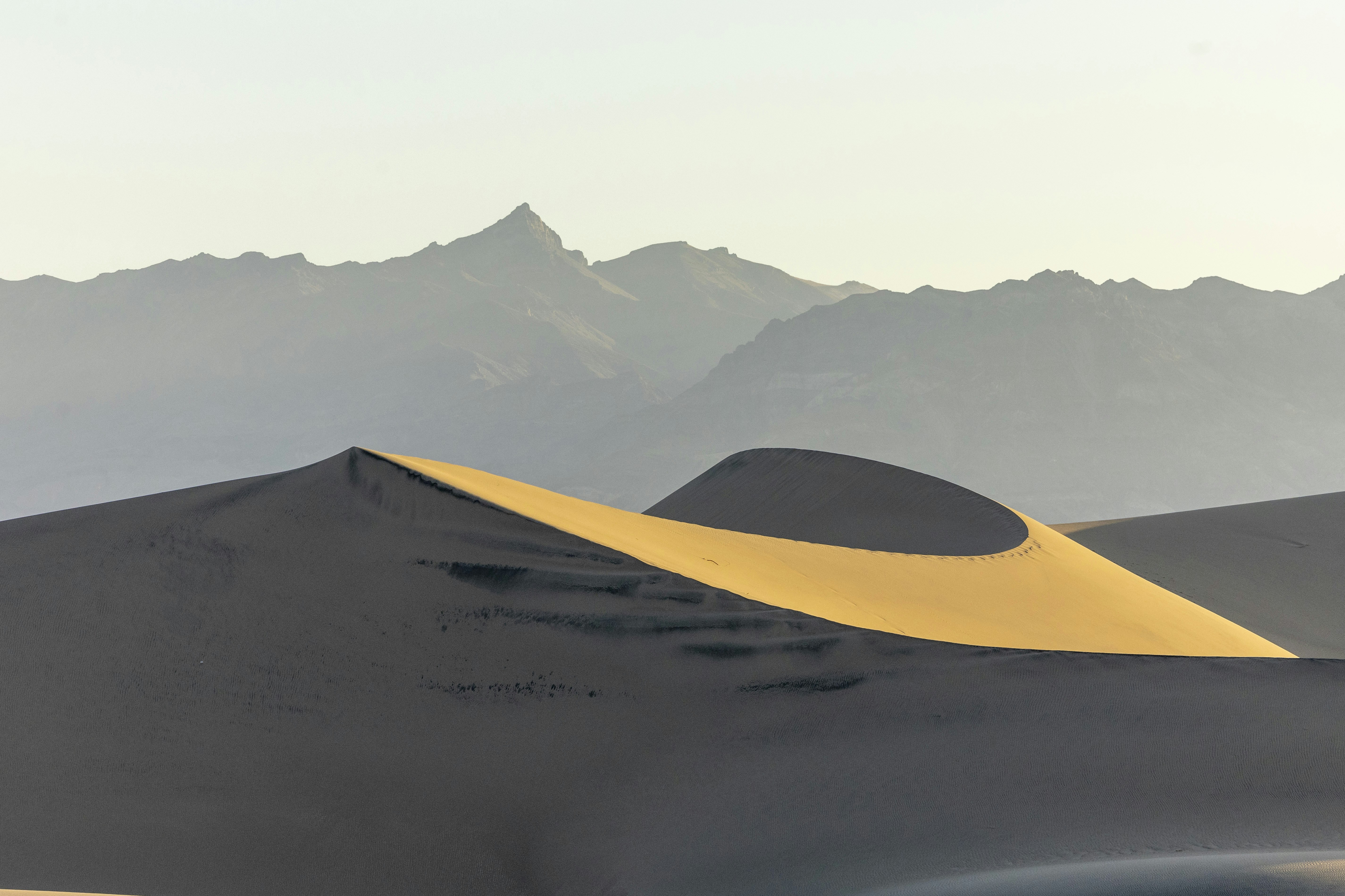 Photograph of a sunlit dune crest winding across a desert with distant, blue-tinted mountains on the horizon. The scene emphasizes light and shadow and expansive negative space.