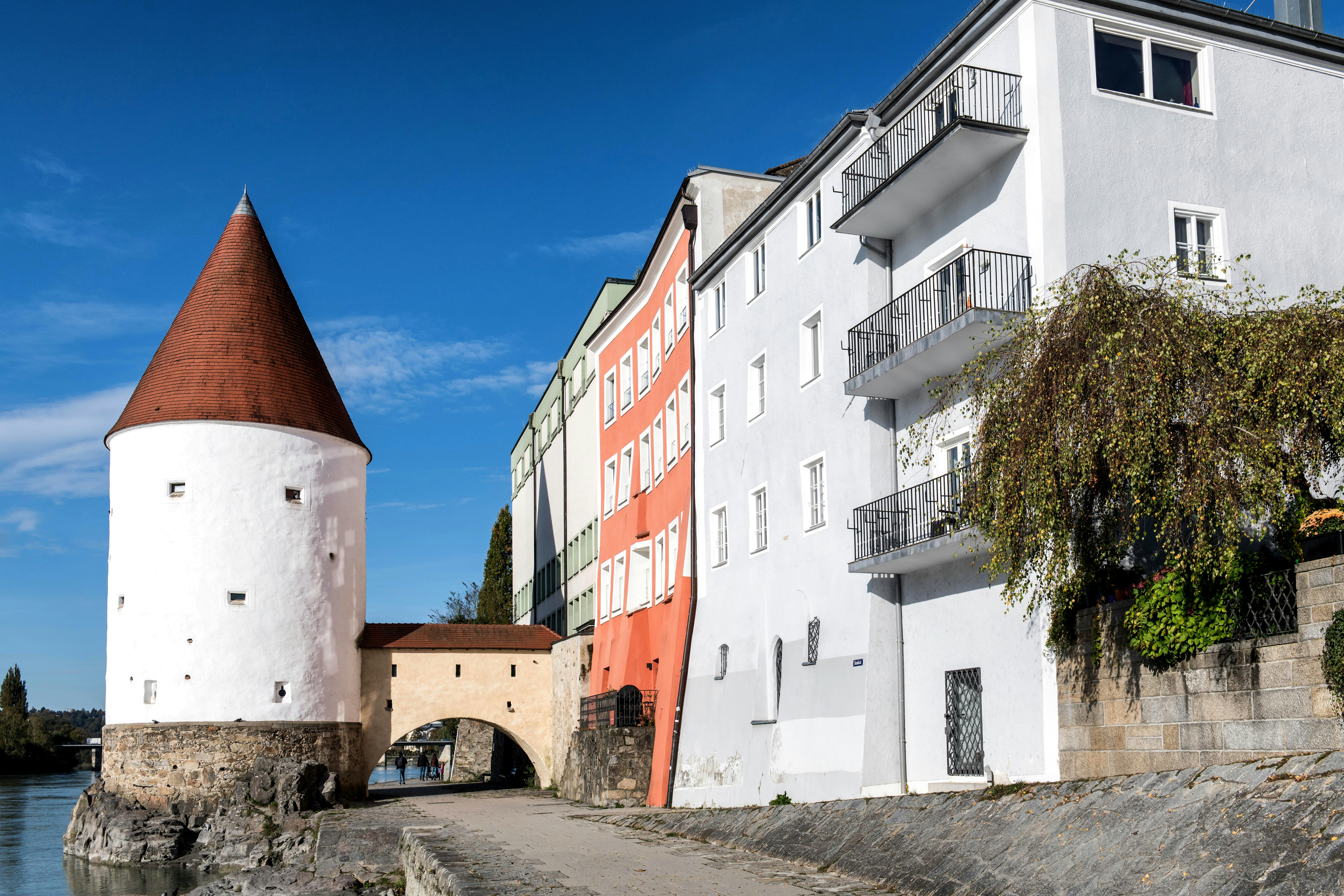 Historic tower with a conical roof adjacent to colorful residential buildings along a waterfront under a clear blue sky.