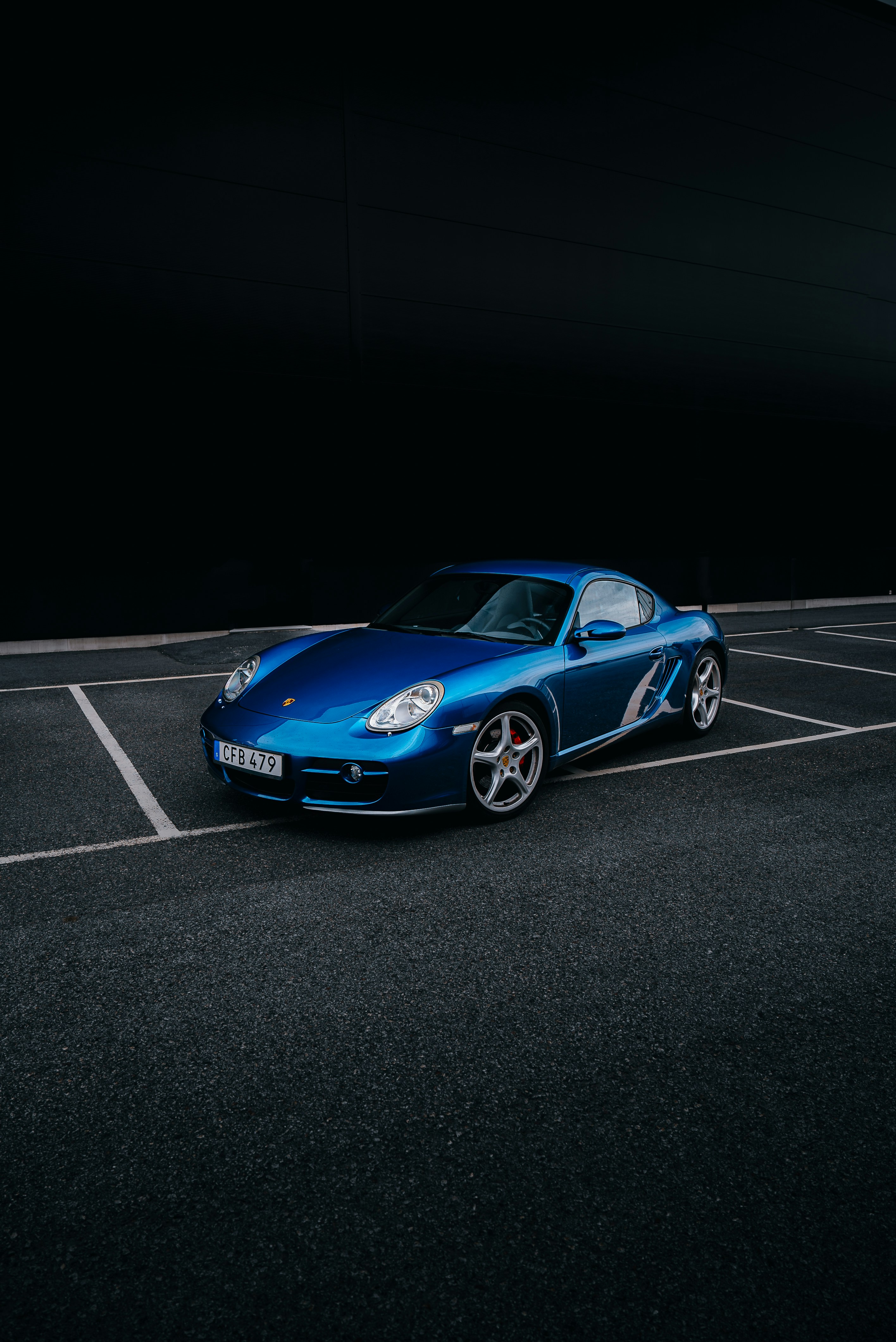 Blue Porsche 911 parked in a nearly empty parking lot beside a dark wall. This high-contrast automotive photograph emphasizes the car's sculpted lines and vivid blue finish.