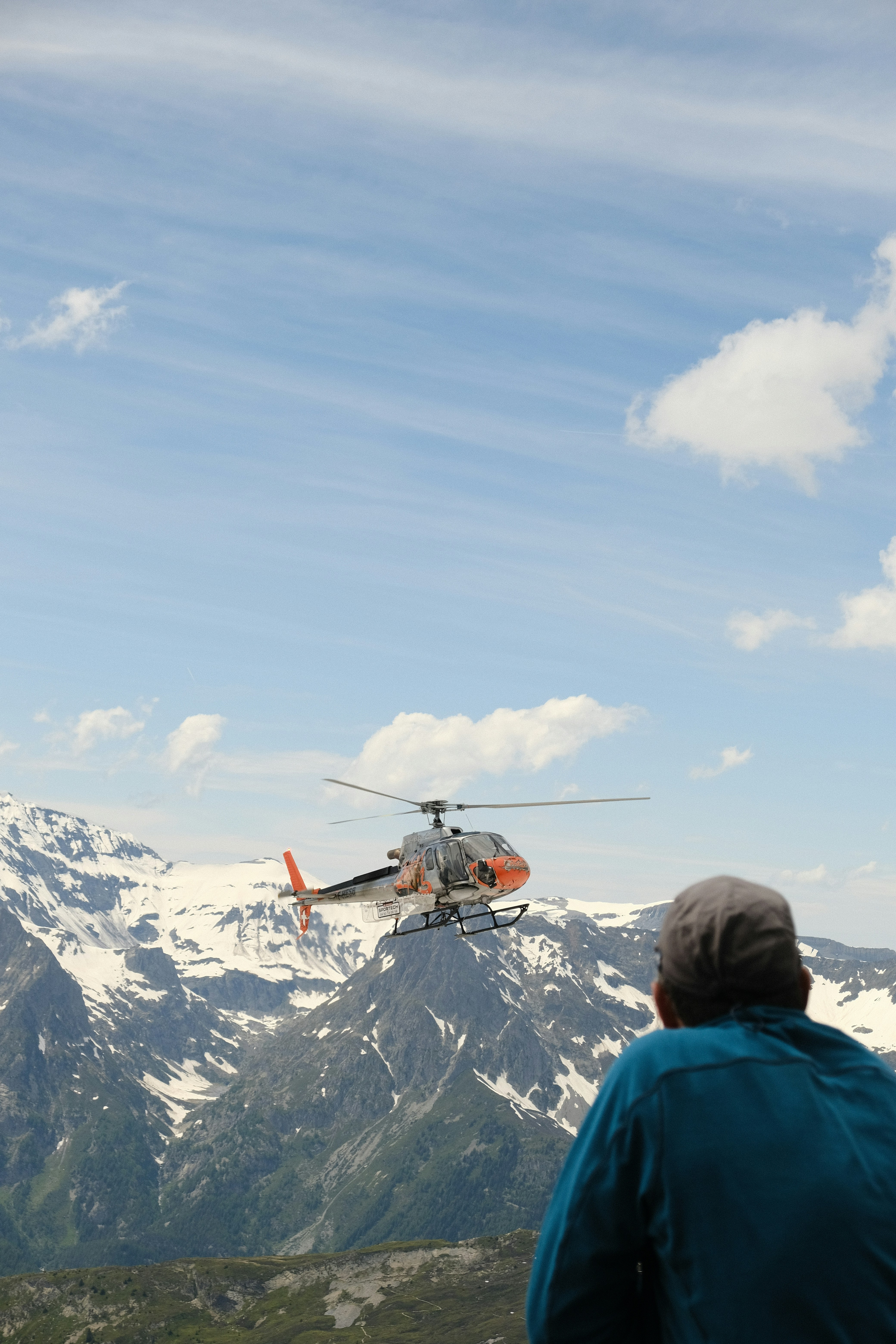 Orange helicopter hovering above serrated Alpine ridges under a crisp blue sky