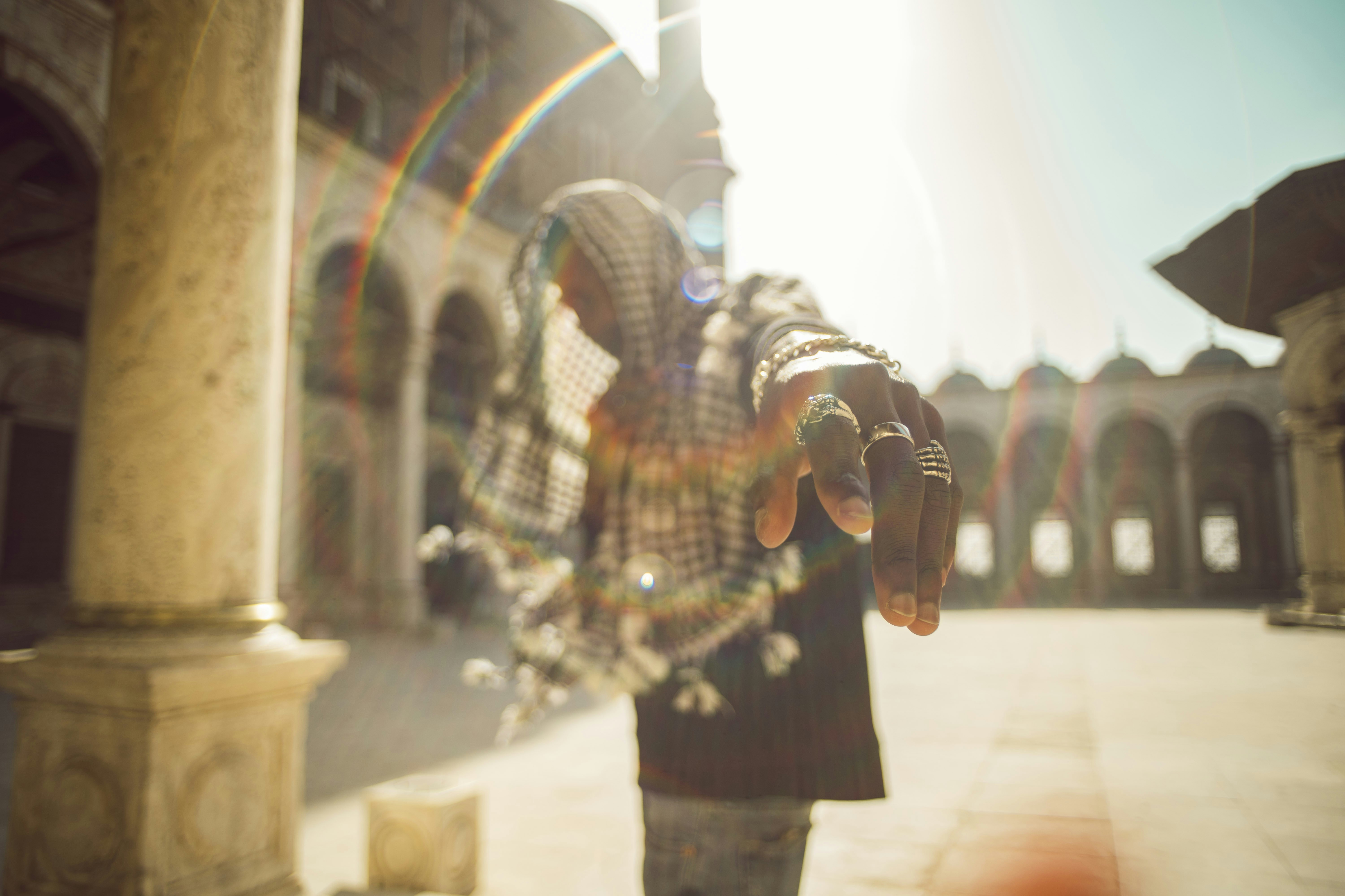 Outstretched Hand Amid Rainbow Flares in a Sunlit Courtyard