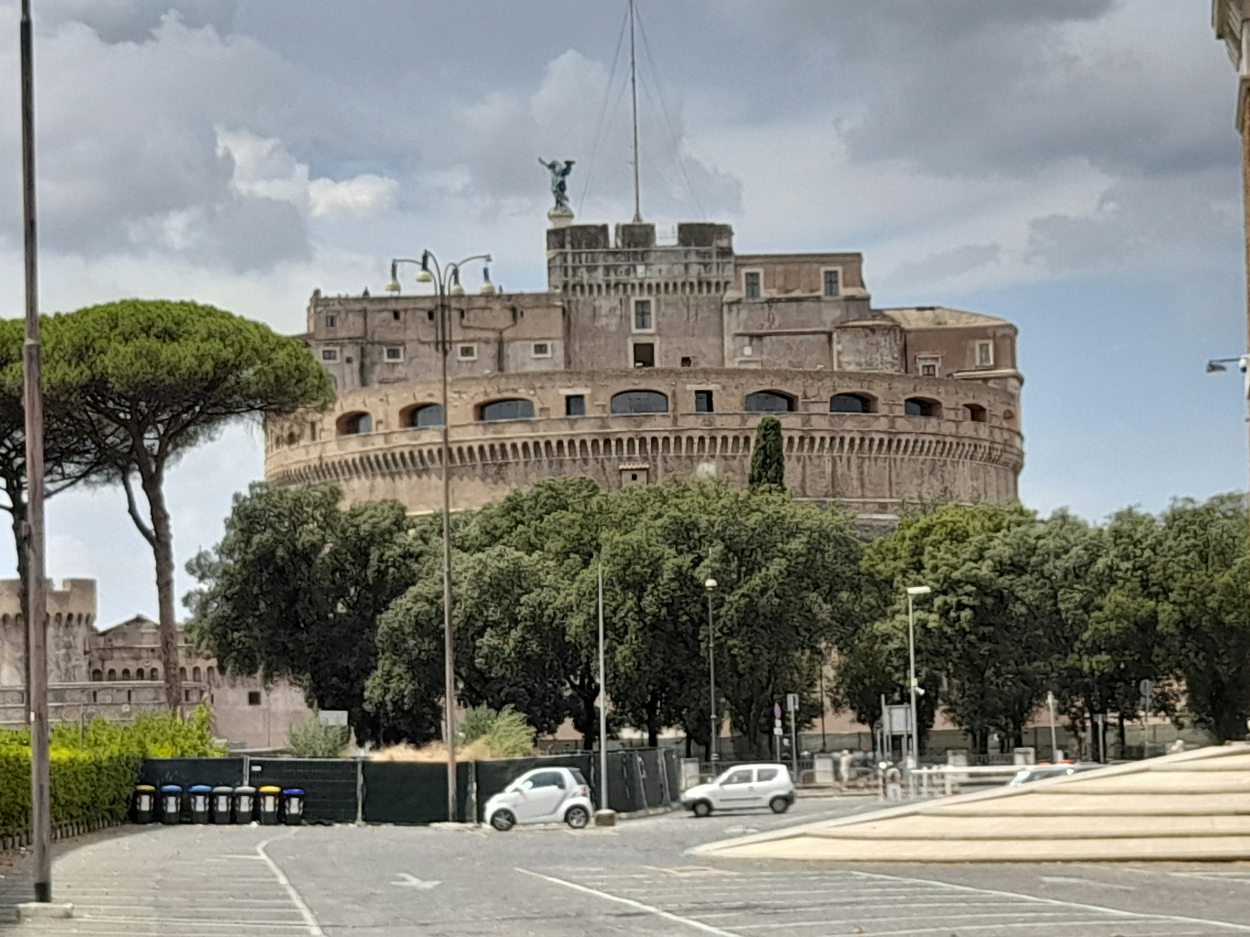 Historic fortress with towering walls under a cloudy sky, surrounded by lush greenery and modern traffic.