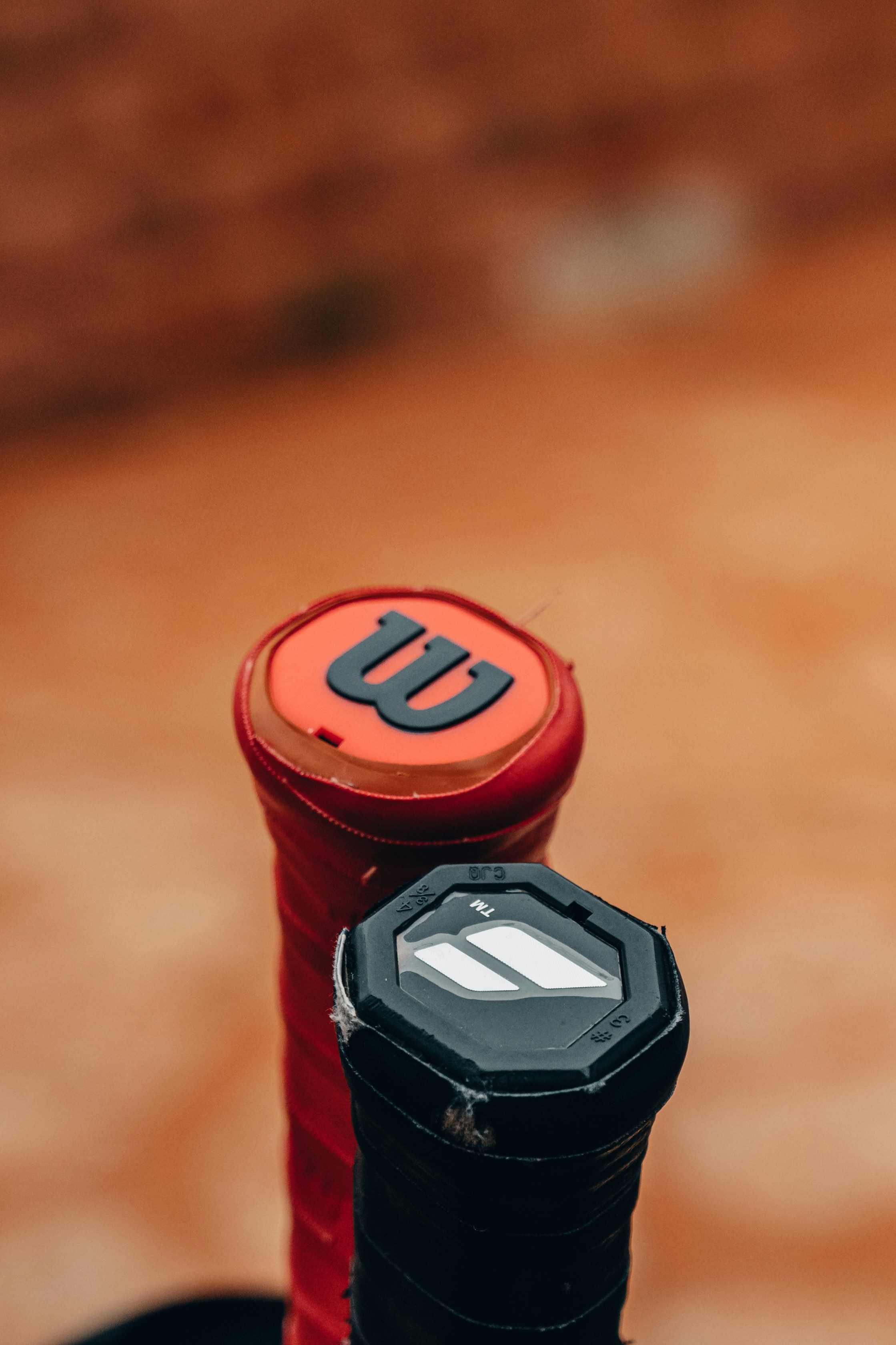 Close-up of two tennis racket grips, one featuring a red Wilson logo and the other a black design, set against a blurred orange background.