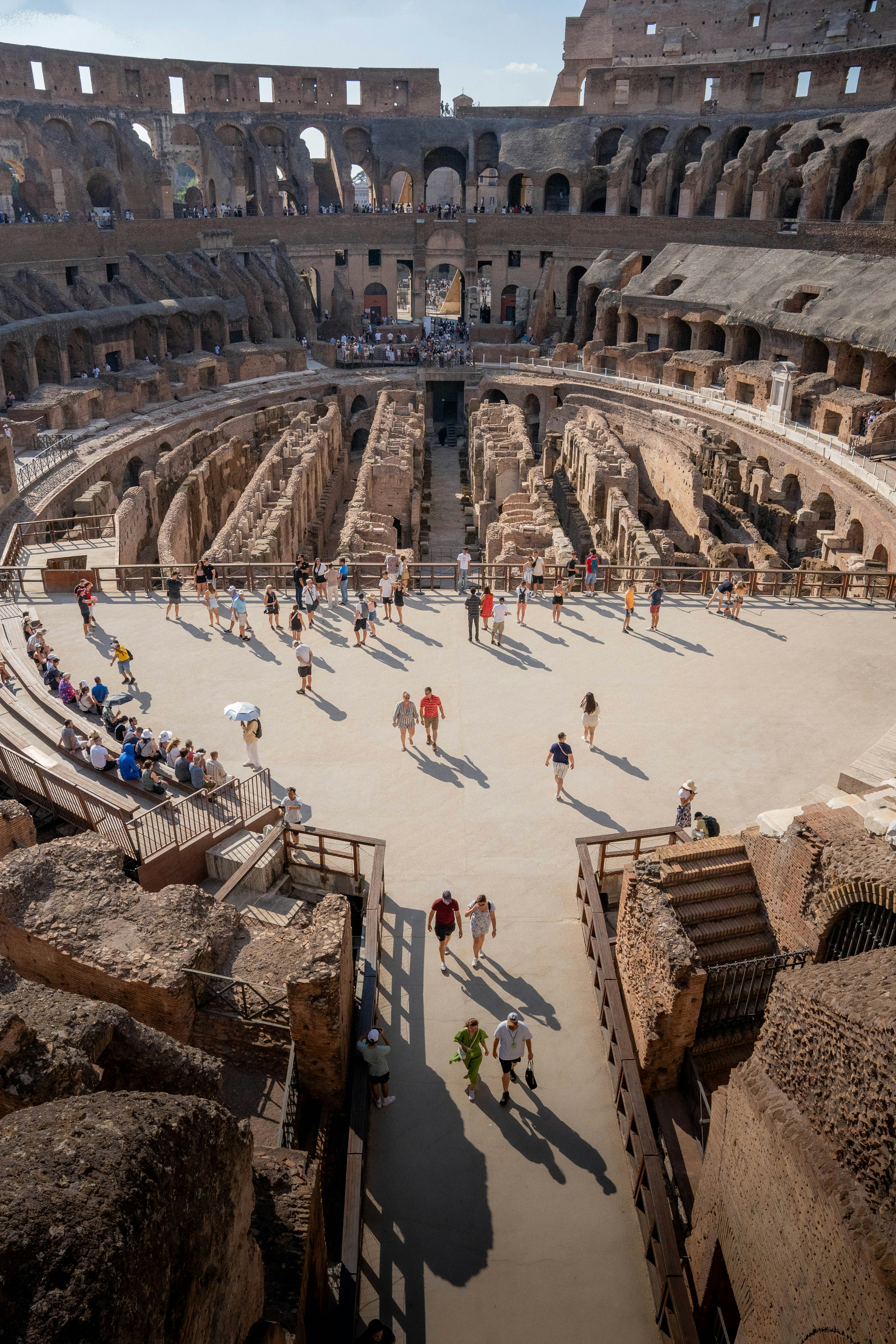 This striking image offers a bird's-eye view of the interior of the Colosseum in Rome, showcasing its ancient grandeur and architectural complexity. The circular arena, once the stage for gladiatorial contests, is surrounded by tiered seating where visitors now stand, soaking in the historical atmosphere. The exposed subterranean structures reveal a labyrinth of passageways and chambers that once held animals and gladiators before they entered the arena. The warm sunlight casts long shadows across the stone floor, highlighting the textures and worn surfaces of the ancient ruins.