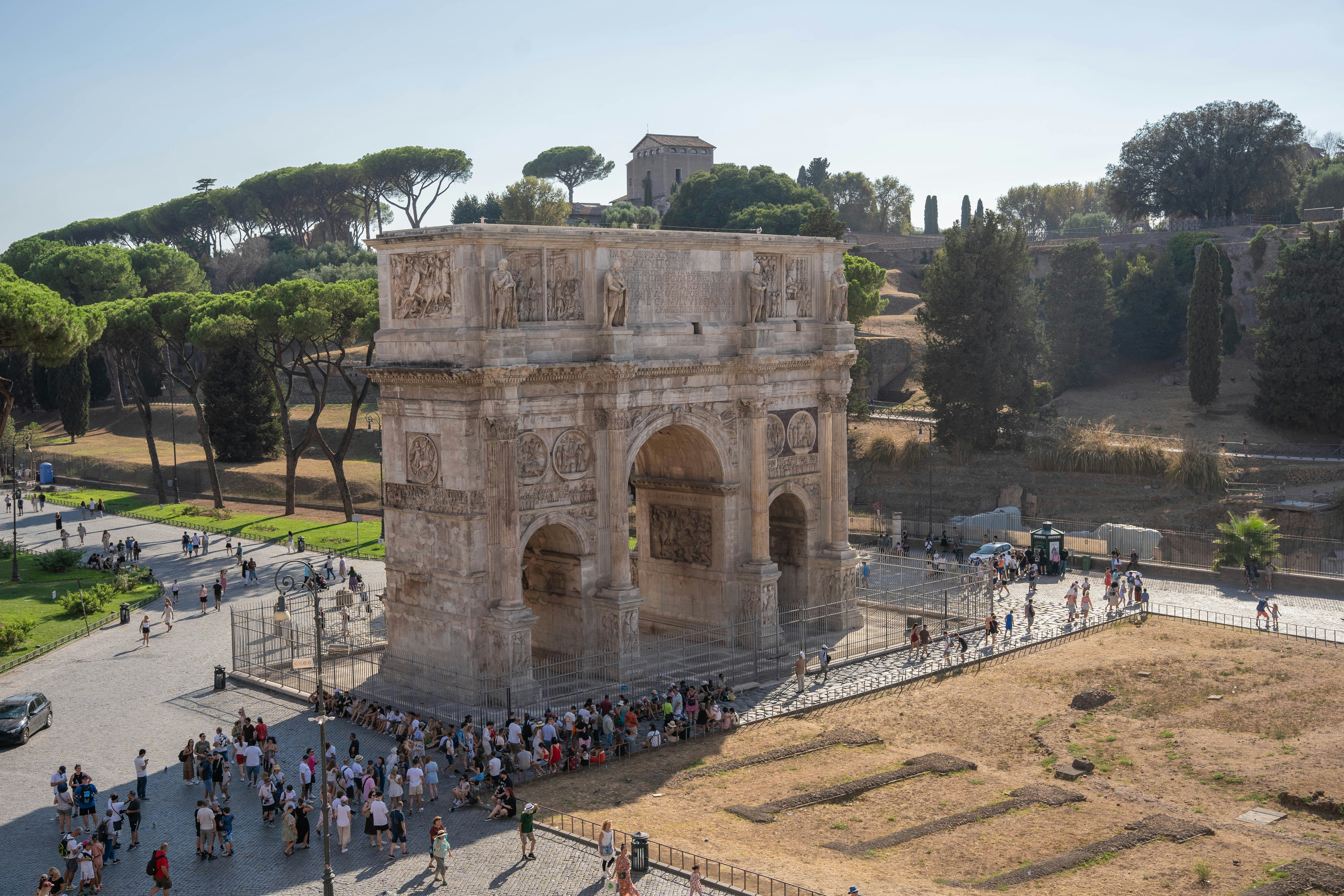 The Arch of Constantine stands prominently near the Colosseum, surrounded by tourists and ancient ruins under a clear sky.