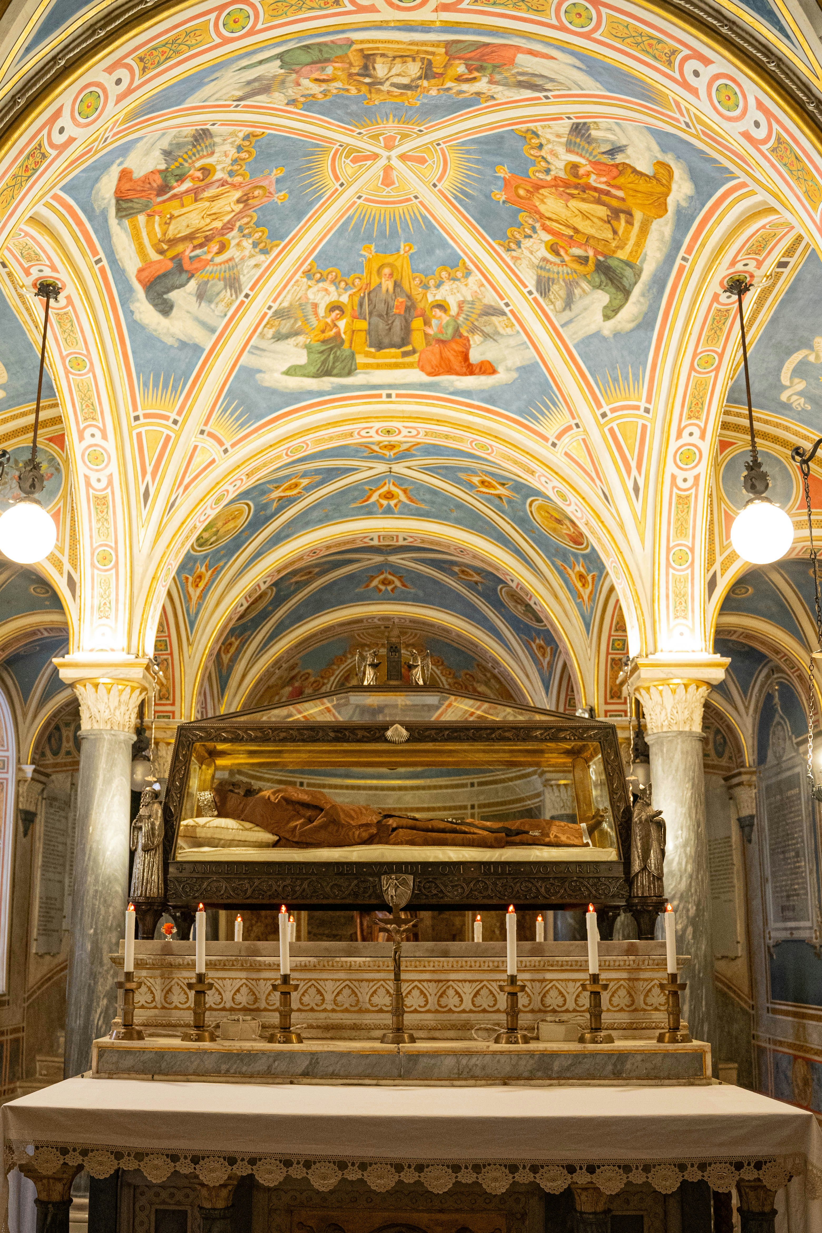 Ornately painted ceiling above an altar, featuring vibrant frescoes and a glass-enclosed relic. Illuminated by soft lighting, it evokes a sense of reverence.