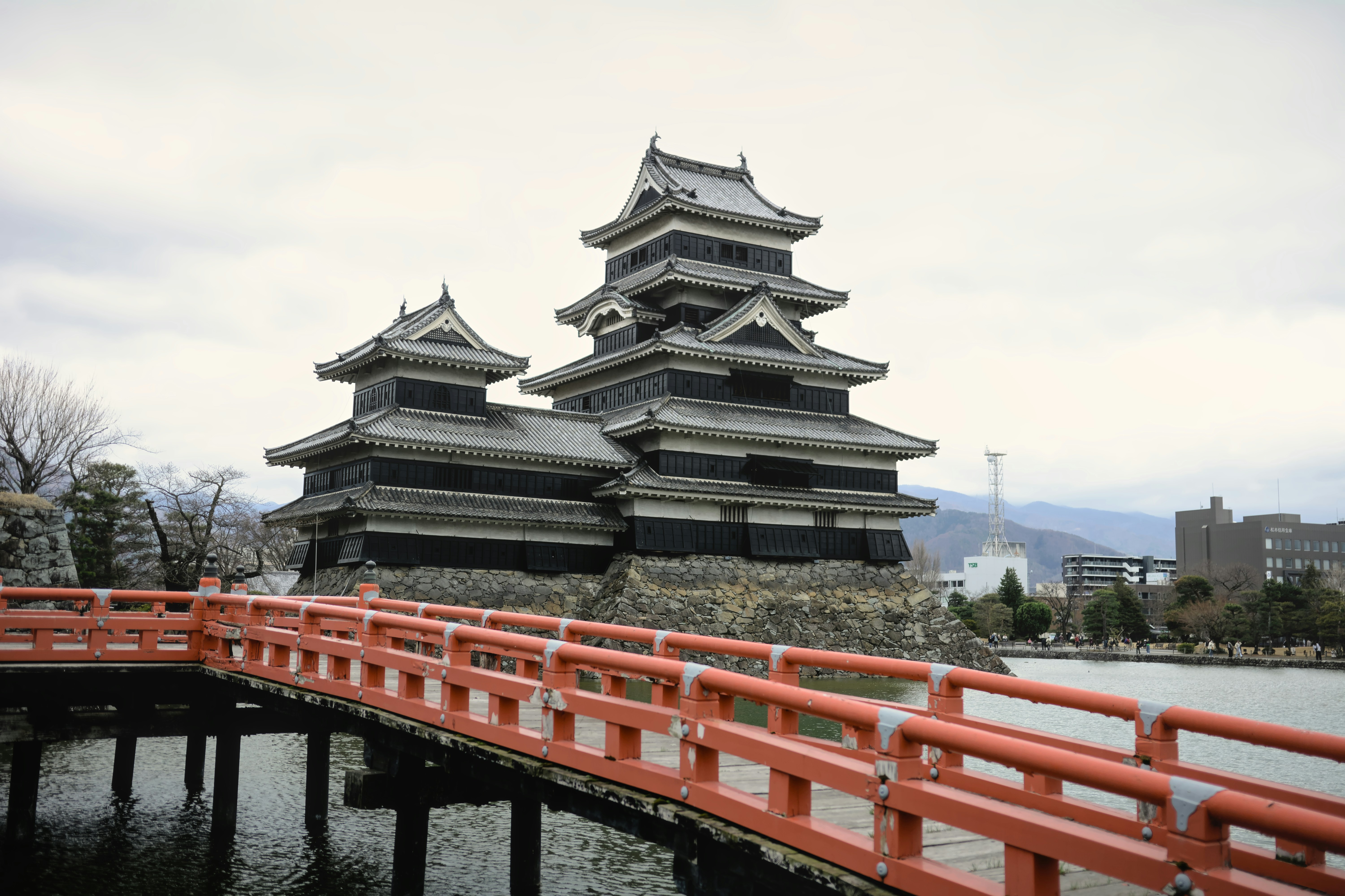Traditional Japanese castle with red bridge crossing a serene moat under a cloudy sky.
