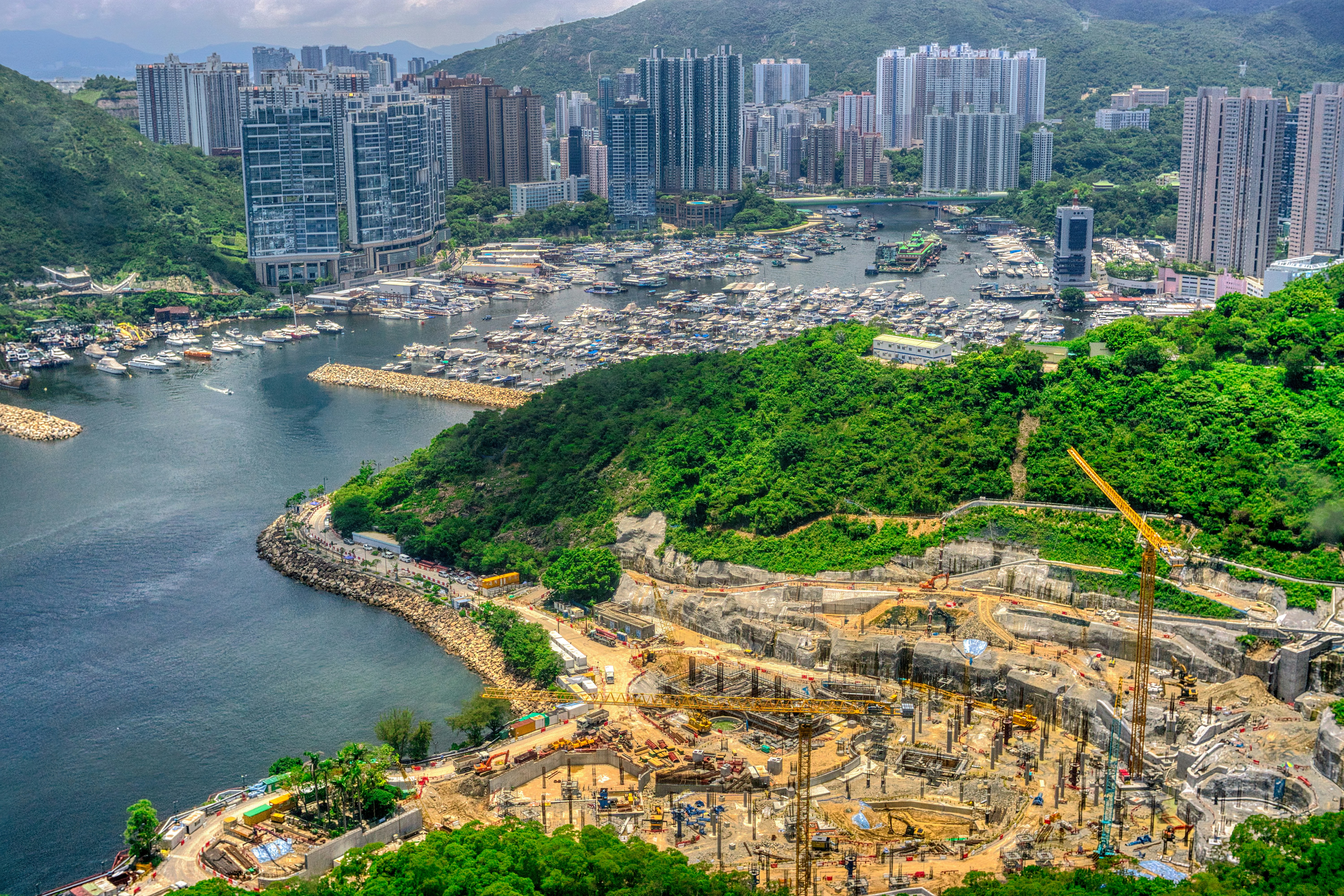 Aerial view of a bustling construction site beside a serene river, framed by lush greenery and towering city buildings.