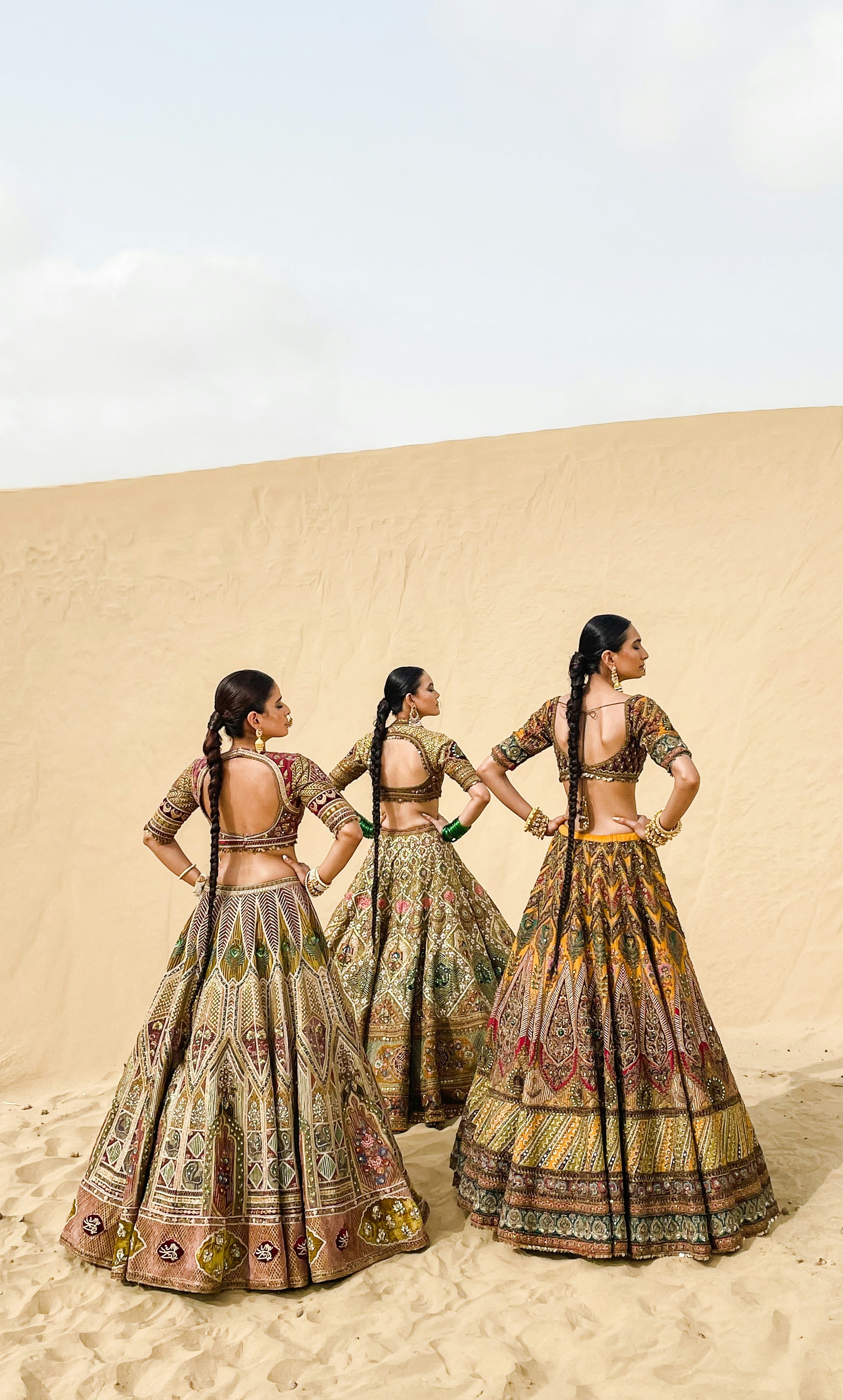 Three women in ornate traditional dresses pose in a sandy desert with a pale dune behind them, highlighting intricate patterns and vibrant colors.