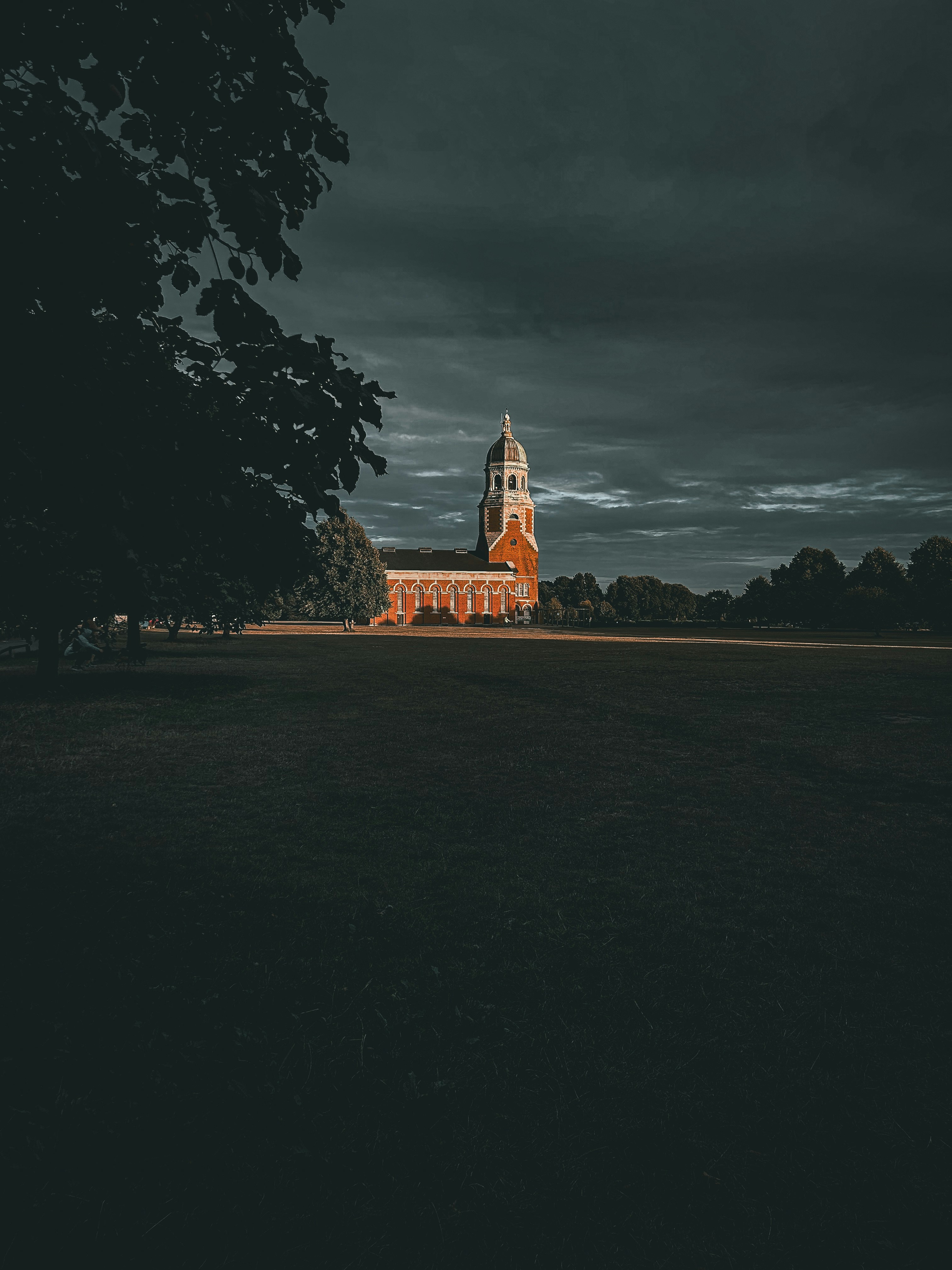 Sunlit red-brick clock tower rises at the center of a spacious park, framed by dark foreground and a moody sky.