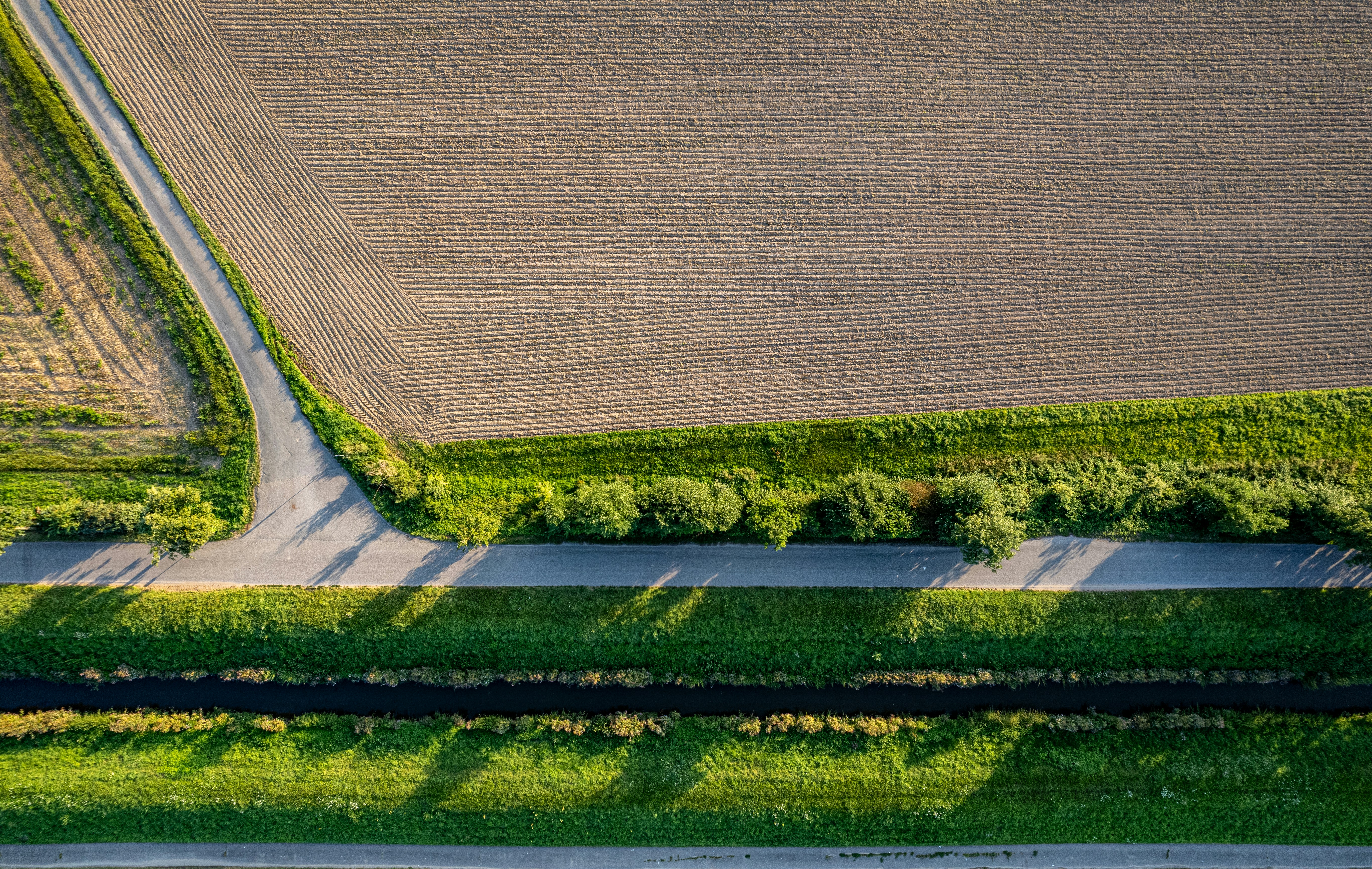 A top-down aerial view of a rural landscape showing a plowed field bordered by a canal and two roads. The field displays neat rows of soil, forming geometric patterns that contrast with the green vegetation along the canal's edge. The canal runs horizontally across the image, dividing the scene into distinct sections. Long shadows from nearby trees and infrastructure add depth and texture to the composition, highlighting the organized nature of the agricultural landscape.