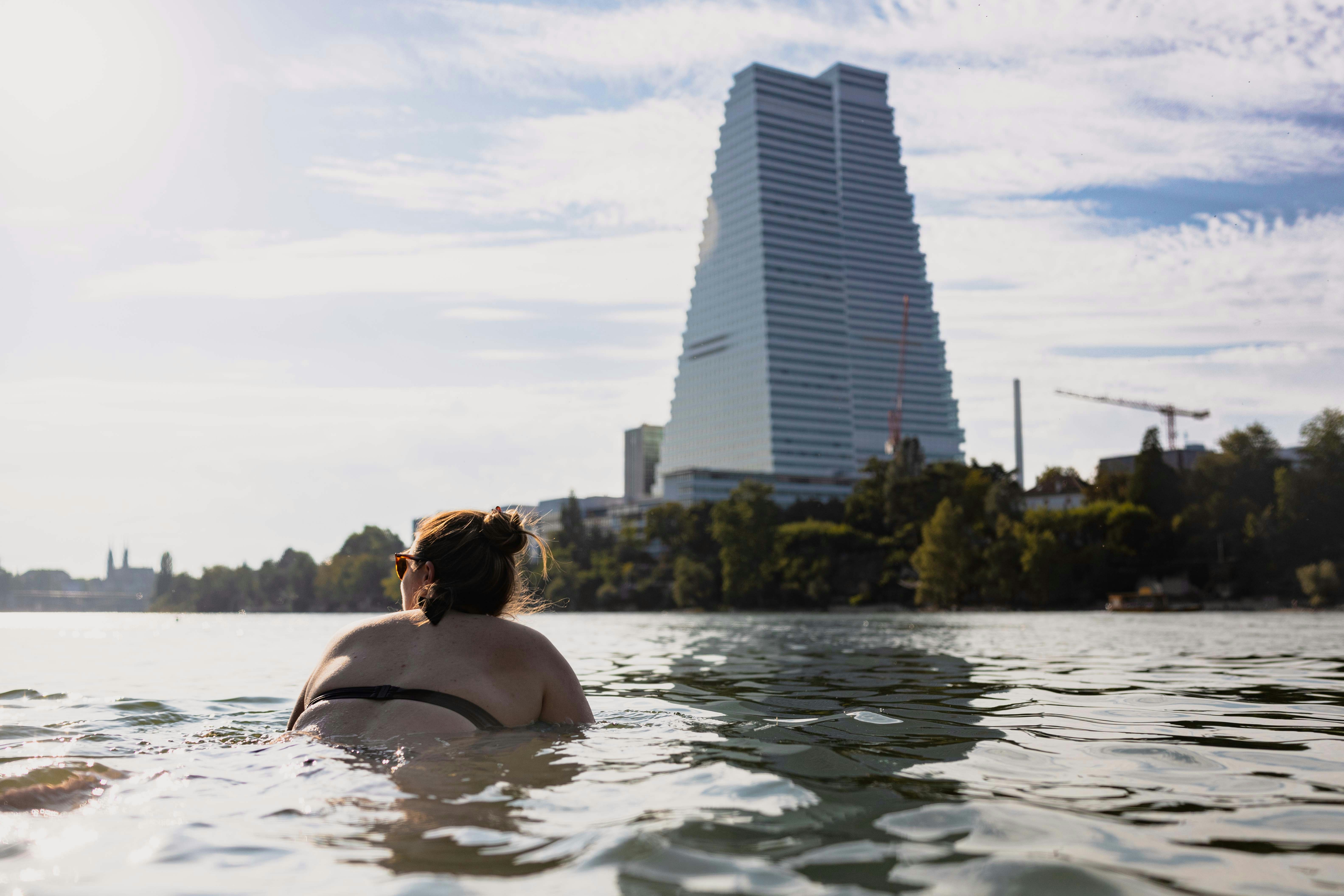 Person swimming in a body of water with a modern skyscraper and trees in the background.