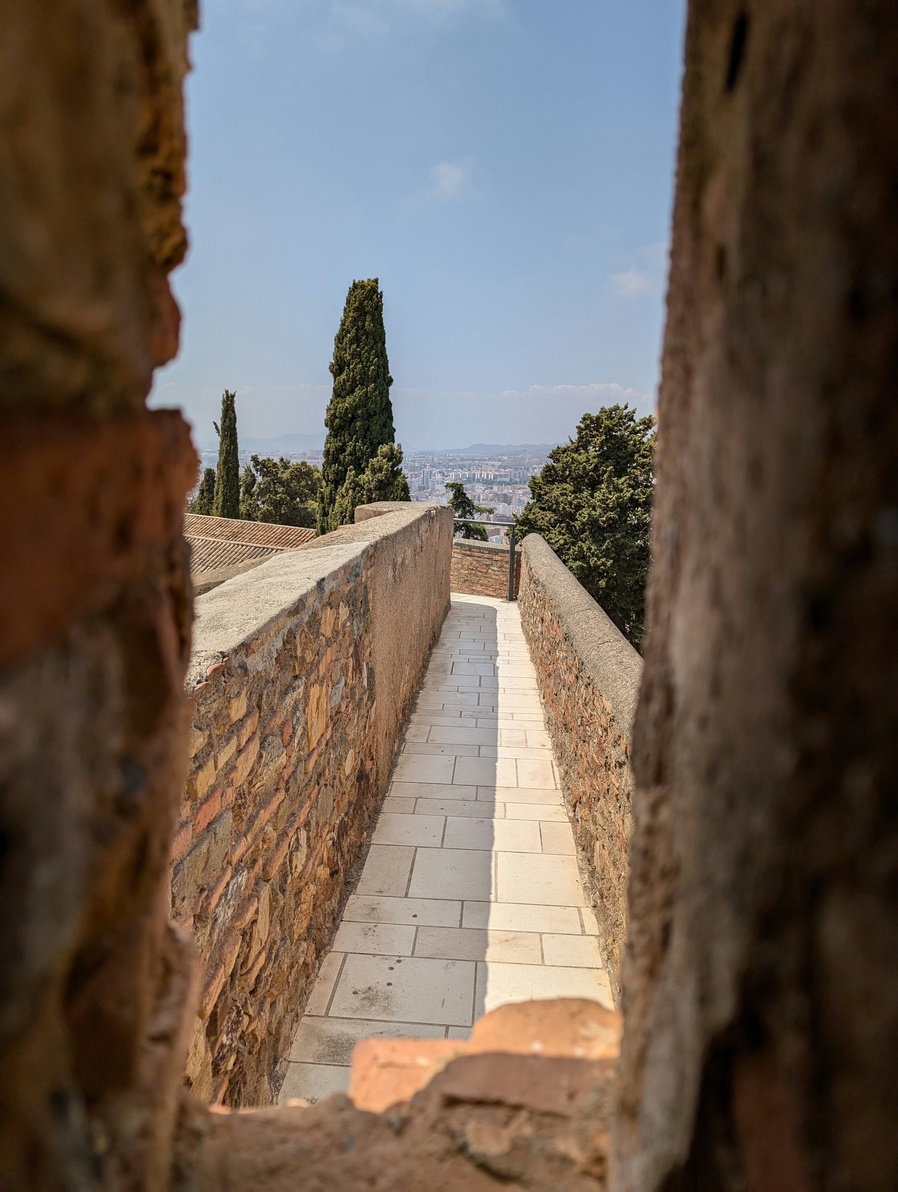 Sunlit stone passage between weathered brick walls leads the eye toward a distant skyline framed by trees and a clear blue sky.