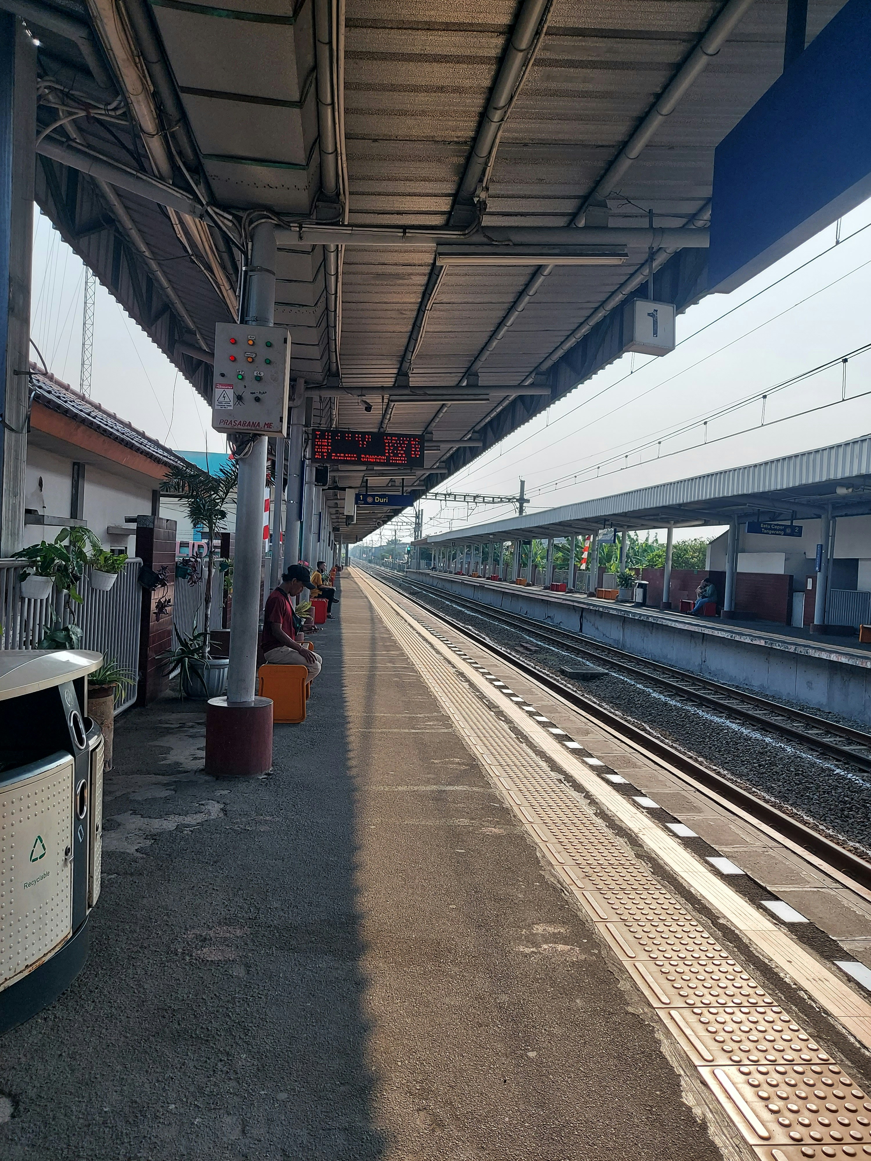 Sunlit railway platform with empty tracks and a digital display board, evoking a sense of anticipation. A few passengers await their trains under the sheltering roof.