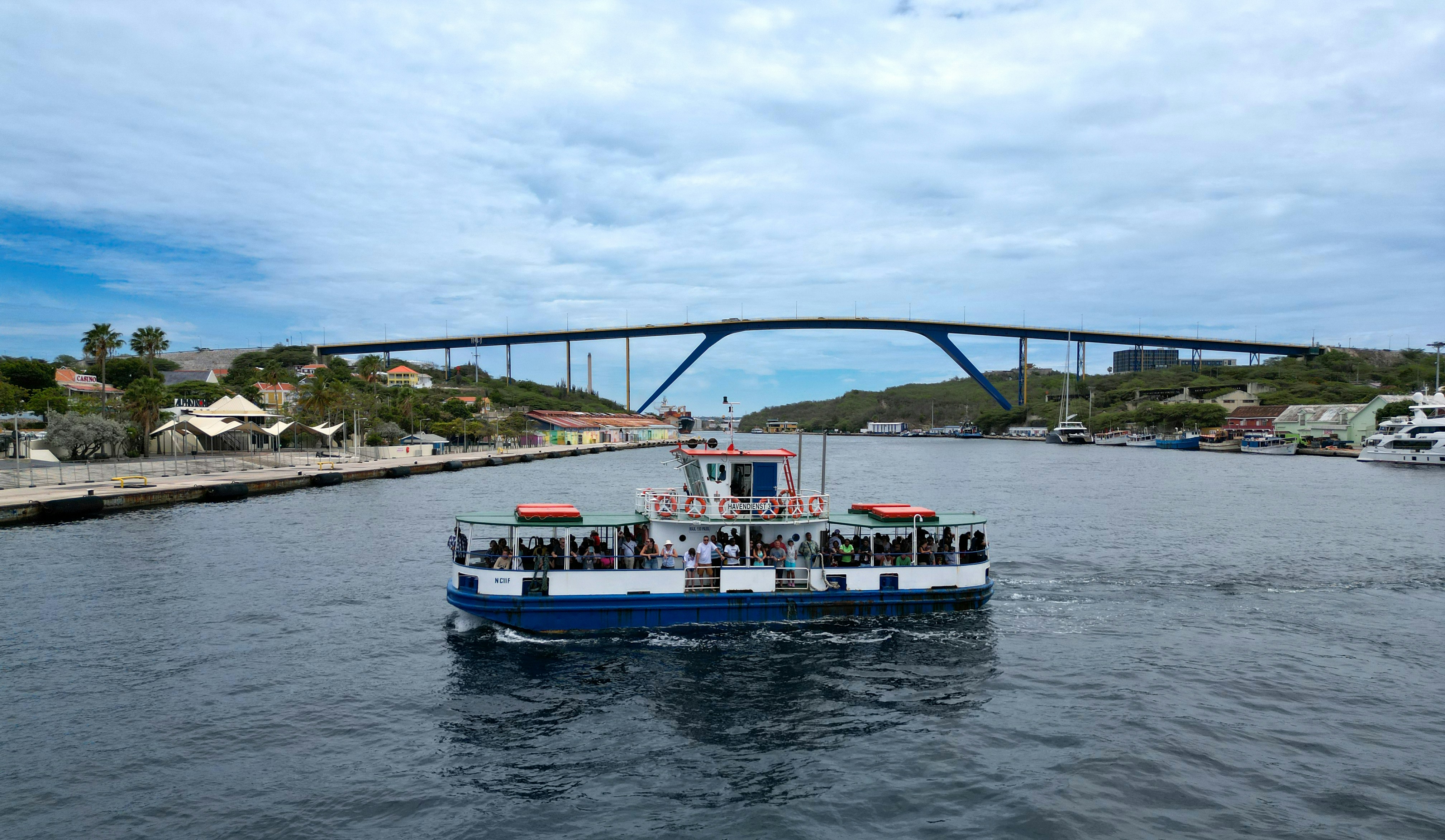A ferry filled with passengers crosses the water under the iconic Queen Juliana Bridge in Willemstad, Curaçao.