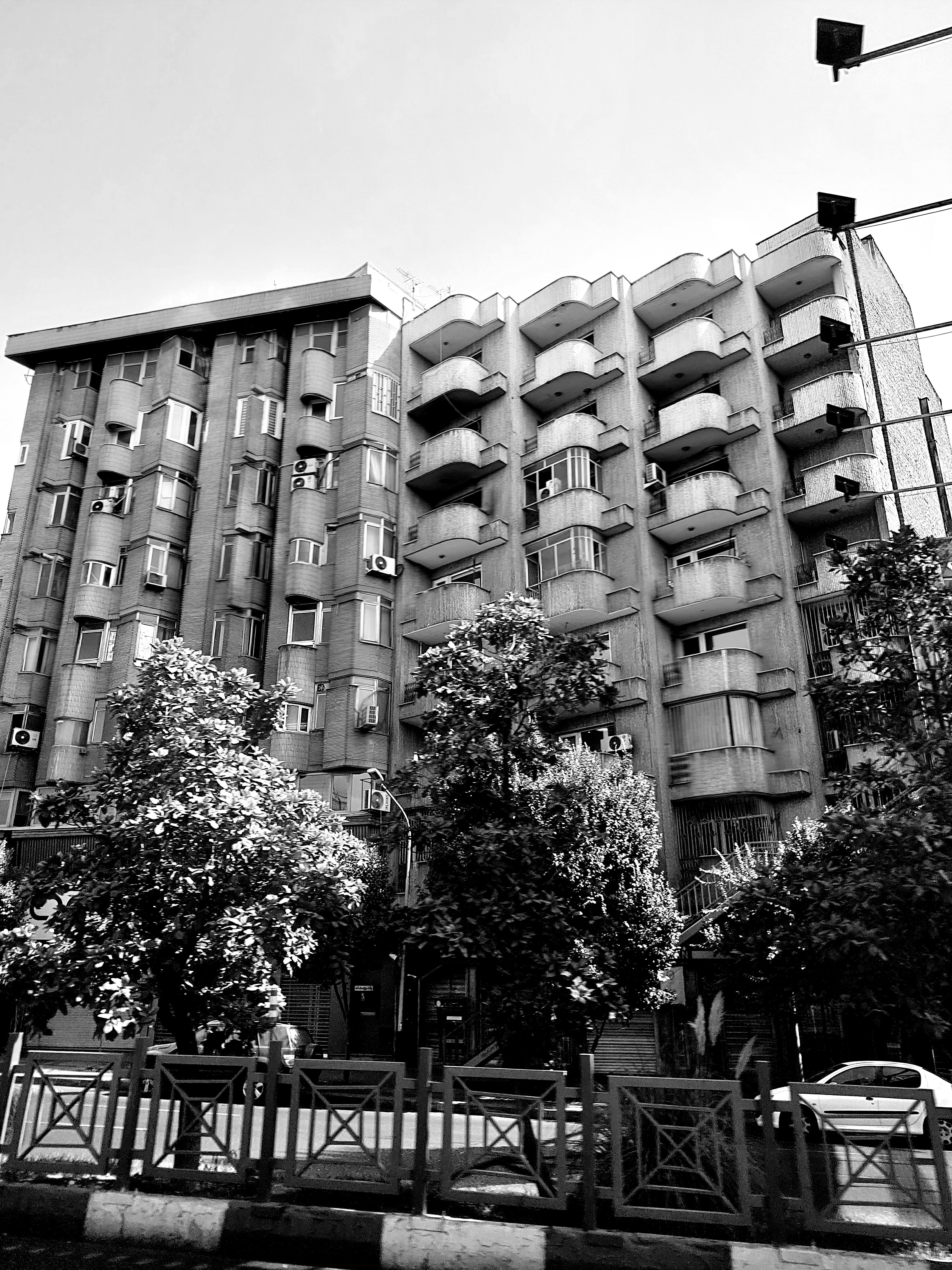 Monochrome street scene featuring a row of apartment buildings with repetitive balconies, framed by trees and a steel railing in the foreground.