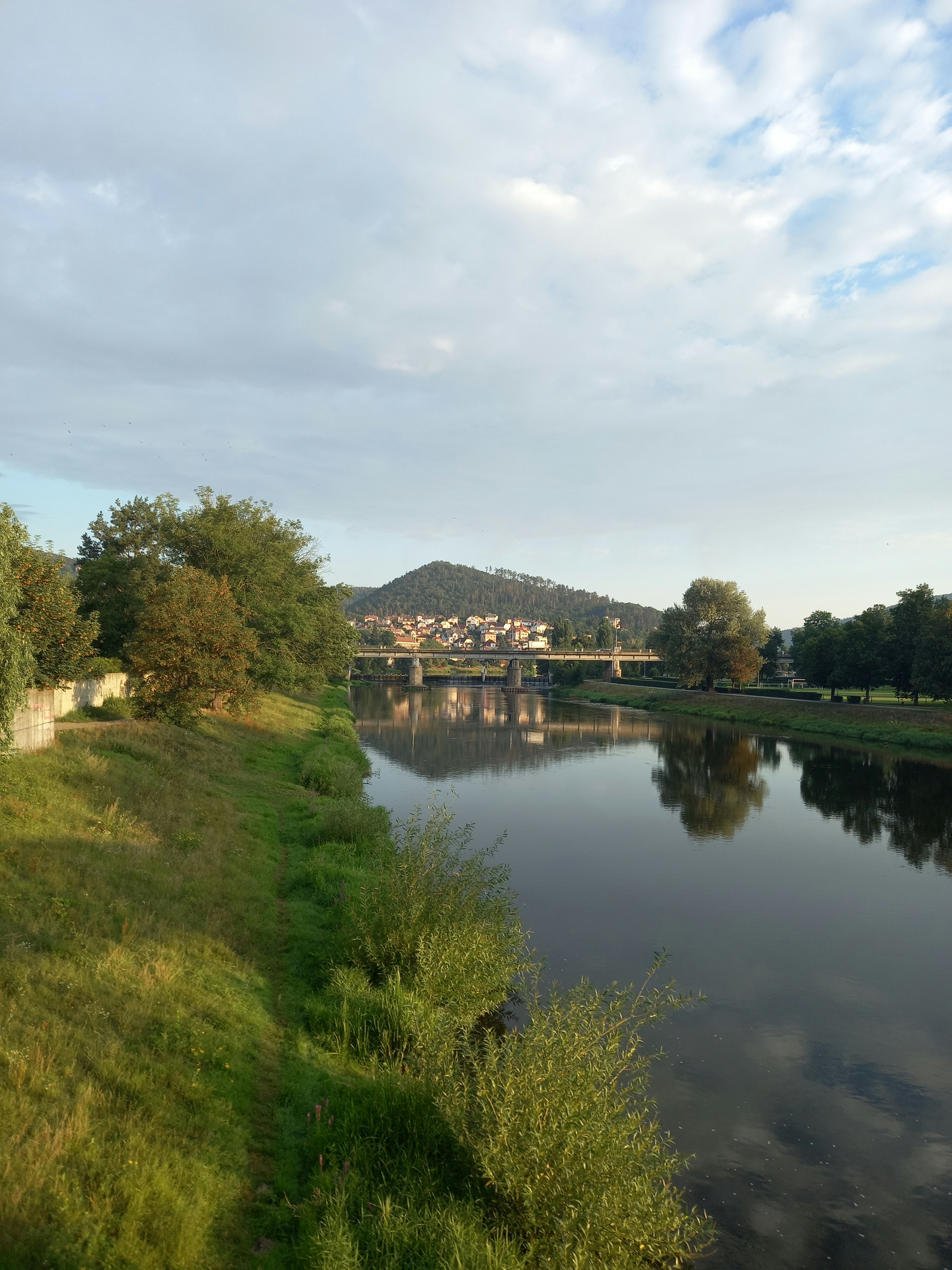 Tranquil river scene with lush greenery and a distant town nestled against a hillside, reflecting in calm waters.