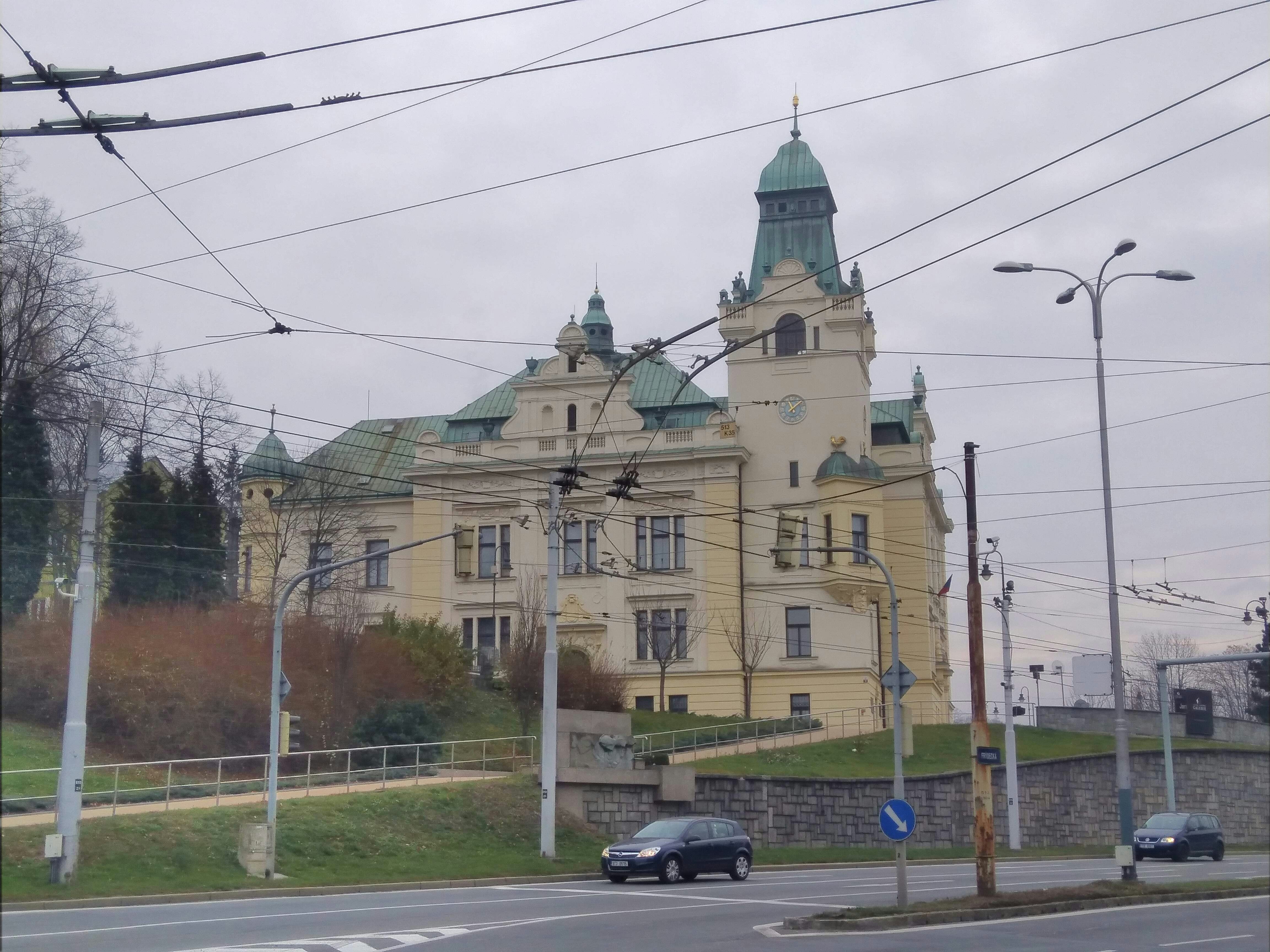 Ornate building with green rooftops stands amid intersecting tram wires and streetlights on a cloudy day.