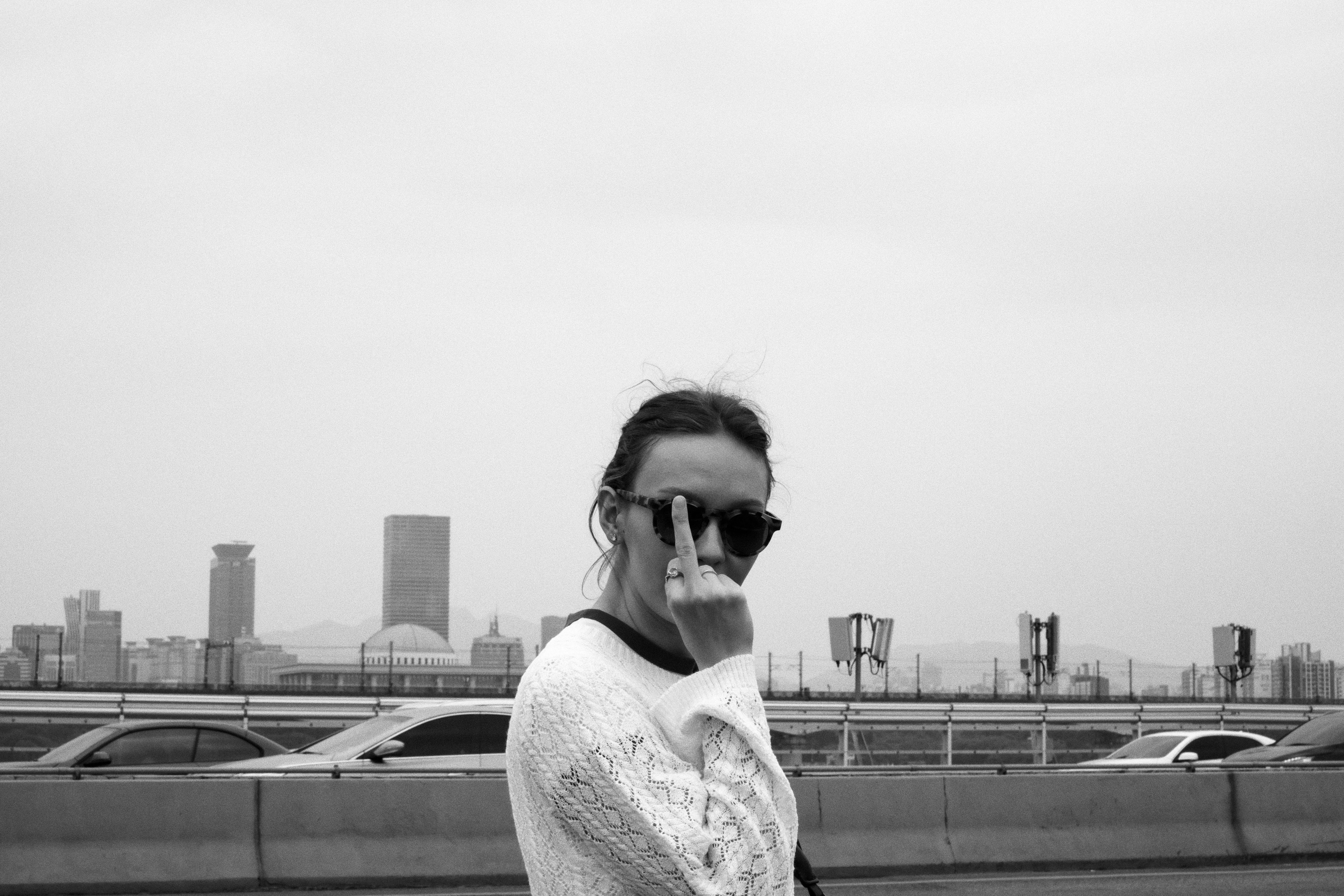 Person in sunglasses and a textured sweater makes a bold gesture with a cityscape backdrop under a soft, overcast sky.