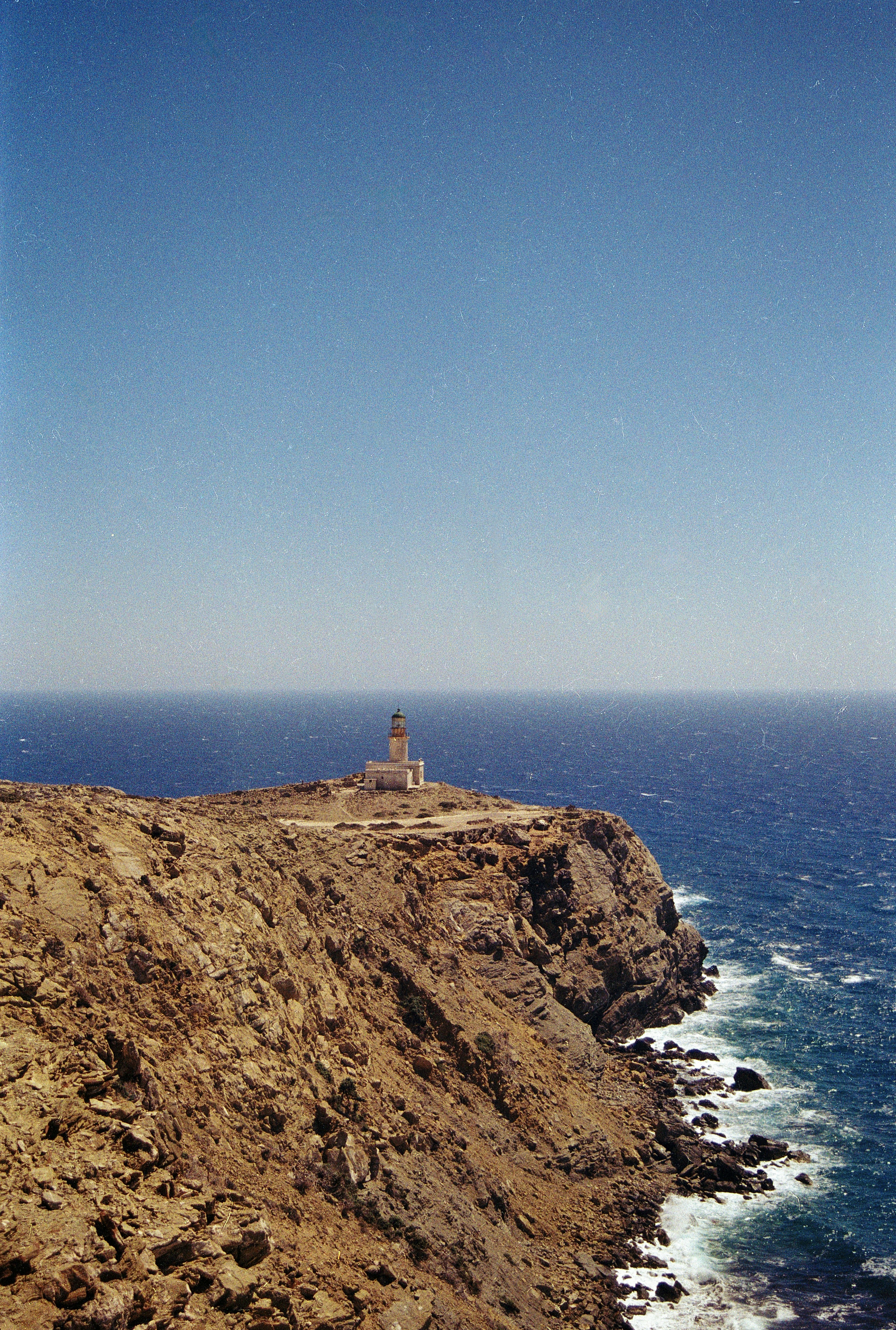 Coastal landscape photograph of a lone lighthouse perched on a rugged promontory above a blue sea.