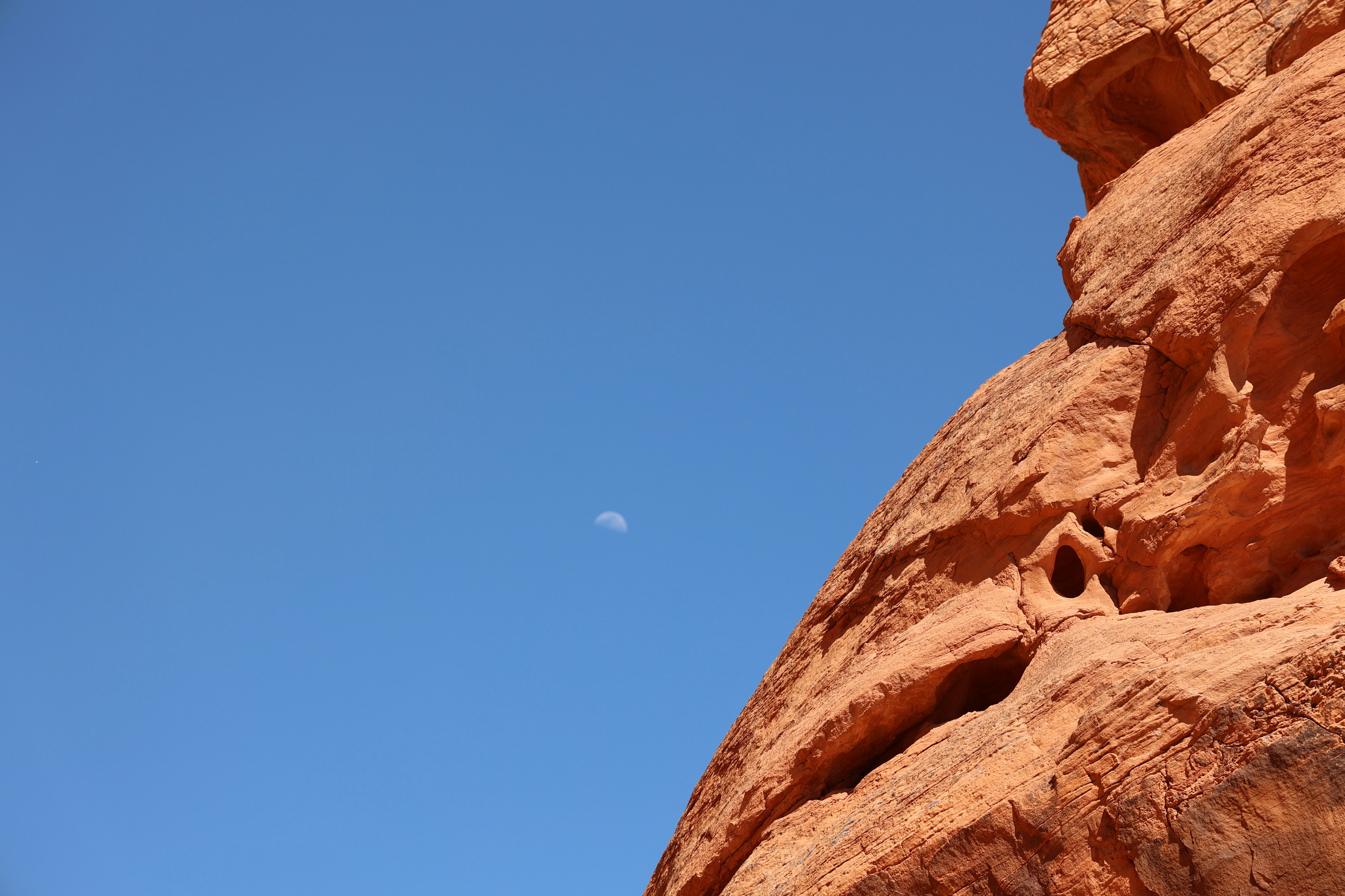 Red rock formation with the moon in a clear blue sky.