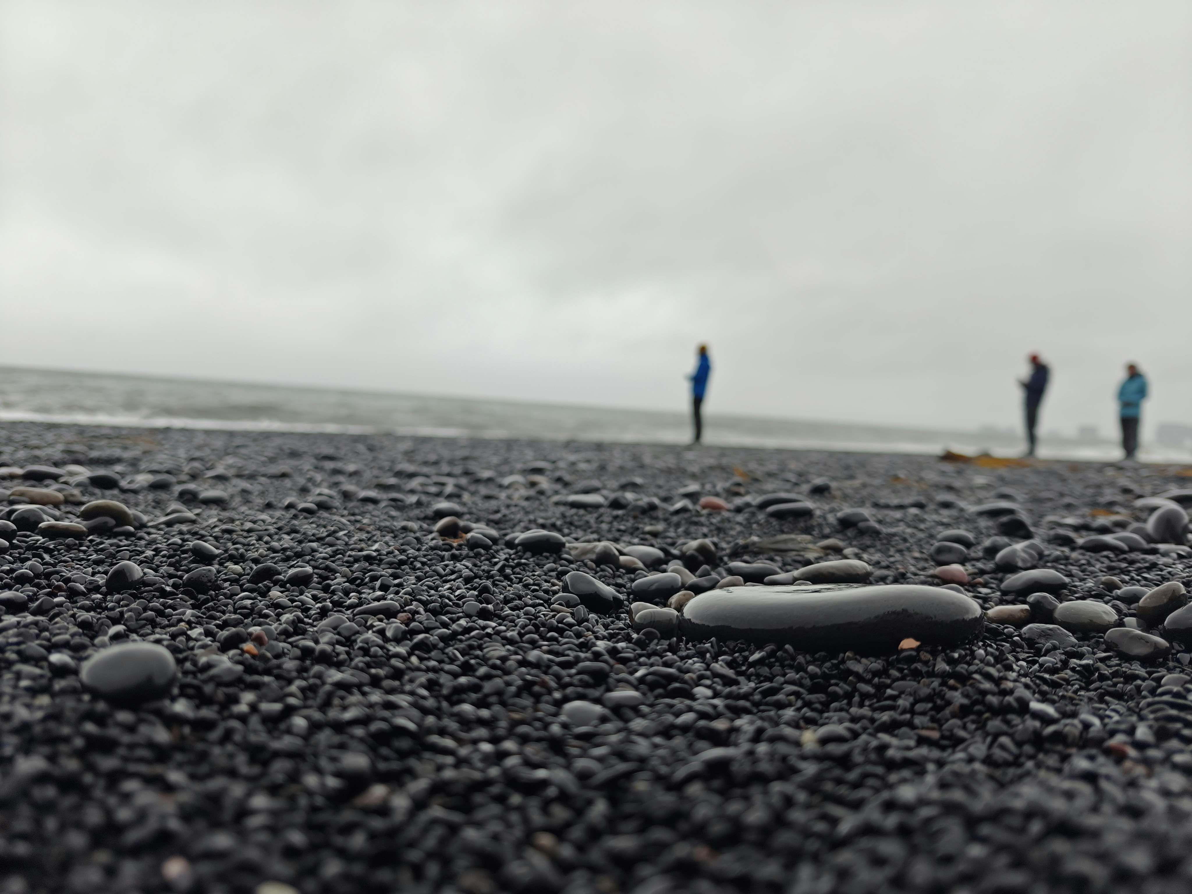 Low-angle shot of a pebble-strewn beach under a gray, overcast sky. Three distant figures stand near the horizon, creating a sense of scale.