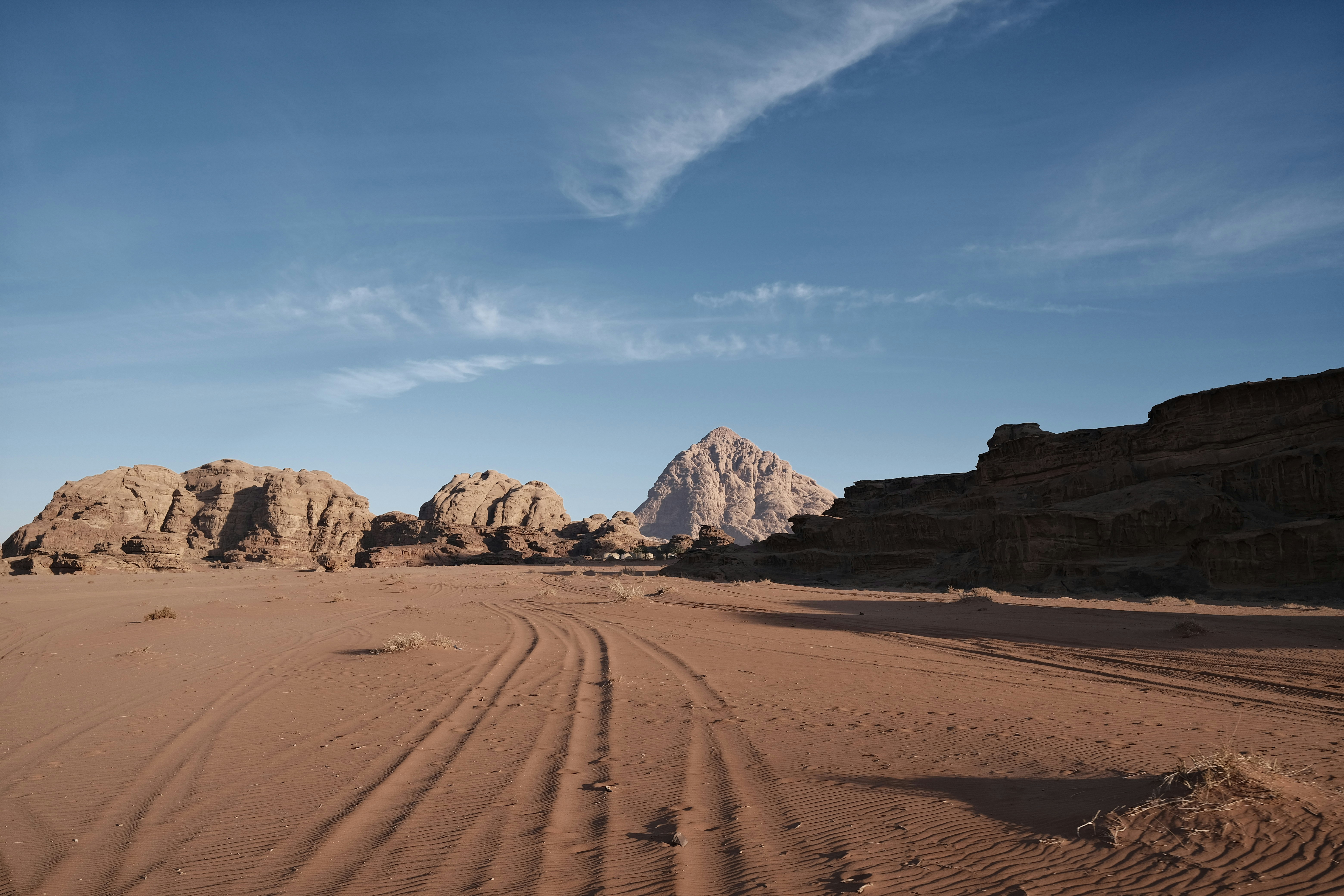 Tire tracks lead through expansive desert sands toward distant rocky formations under a clear blue sky.