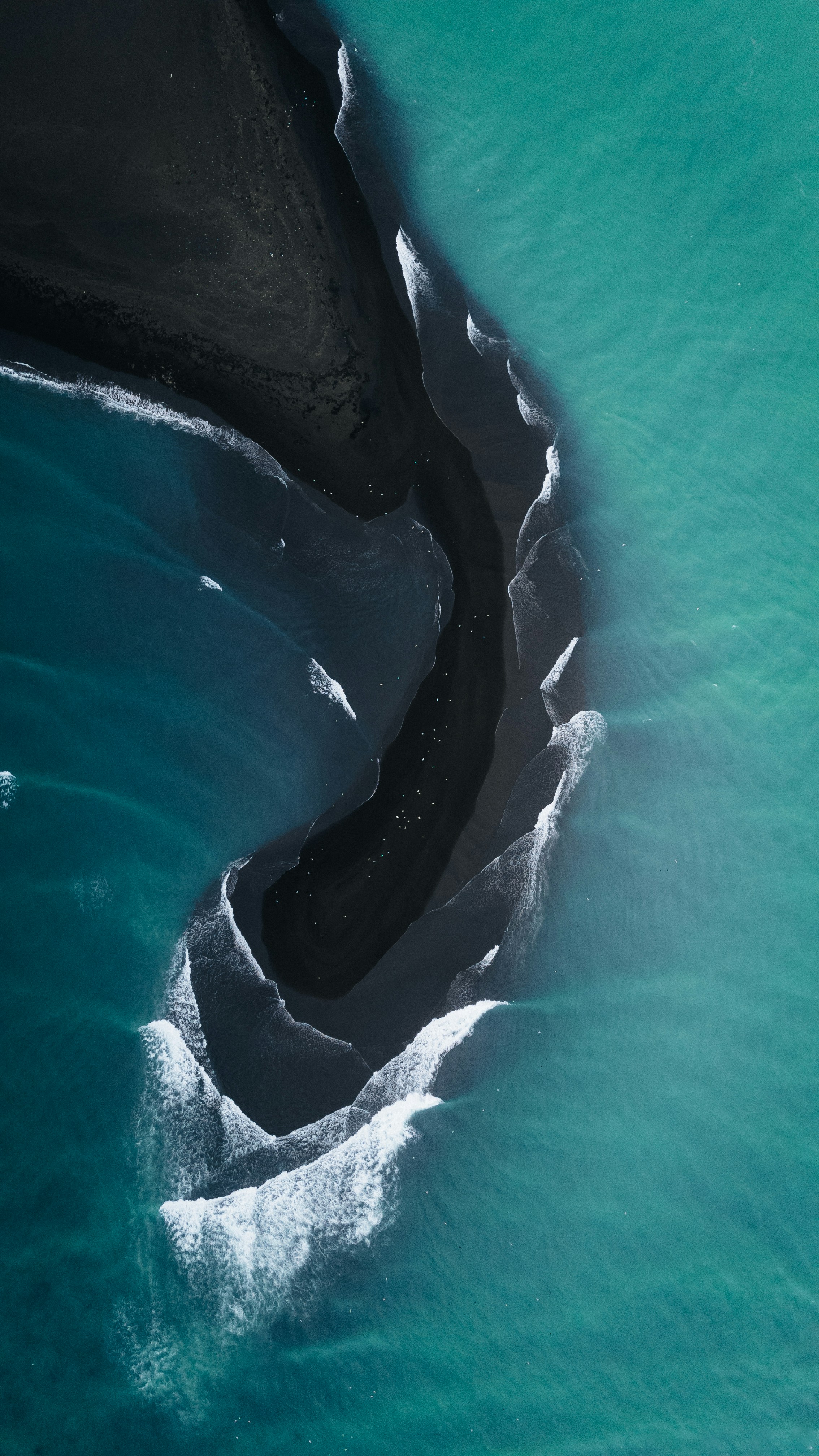 Black sand beach curves against vibrant turquoise waters with gentle white-tipped waves.