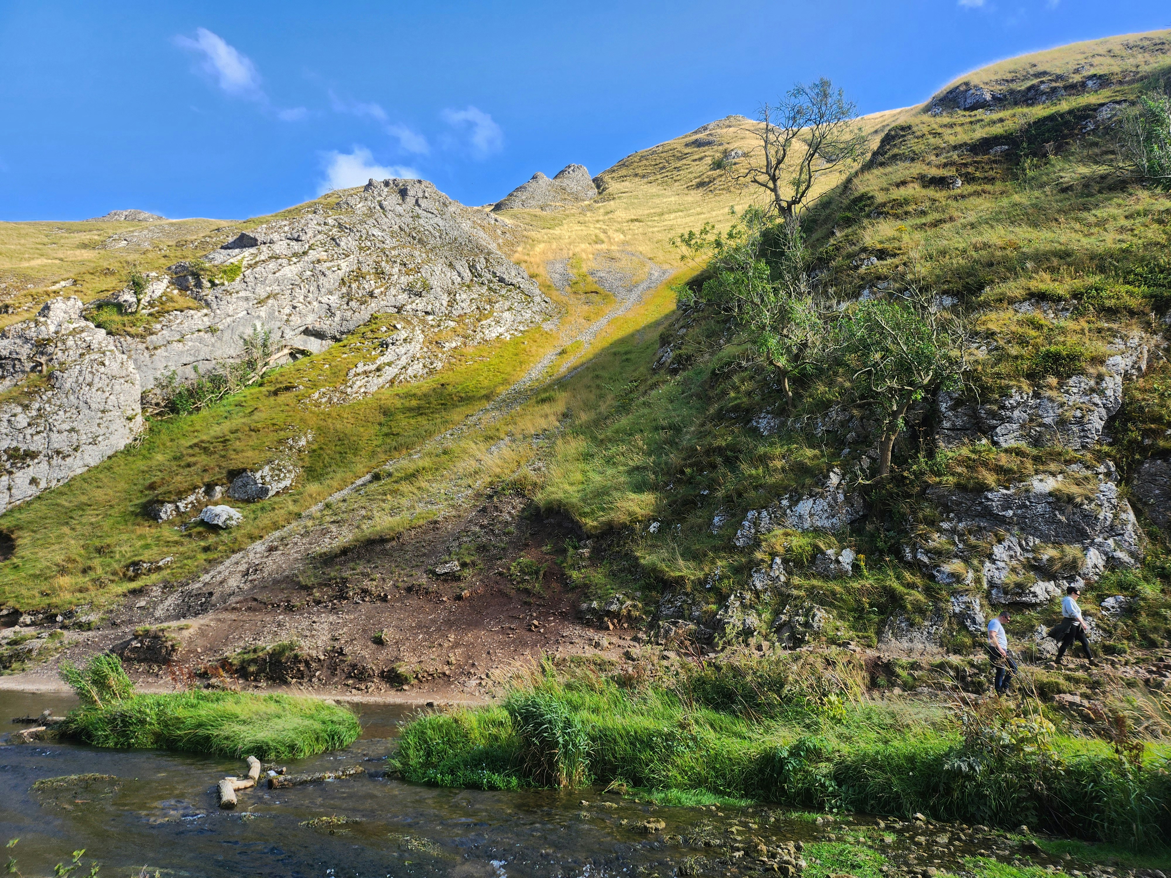 Lush green hillside meets a serene stream, showcasing the beauty of natural landscapes. A couple of hikers explore the terrain, adding a sense of scale.