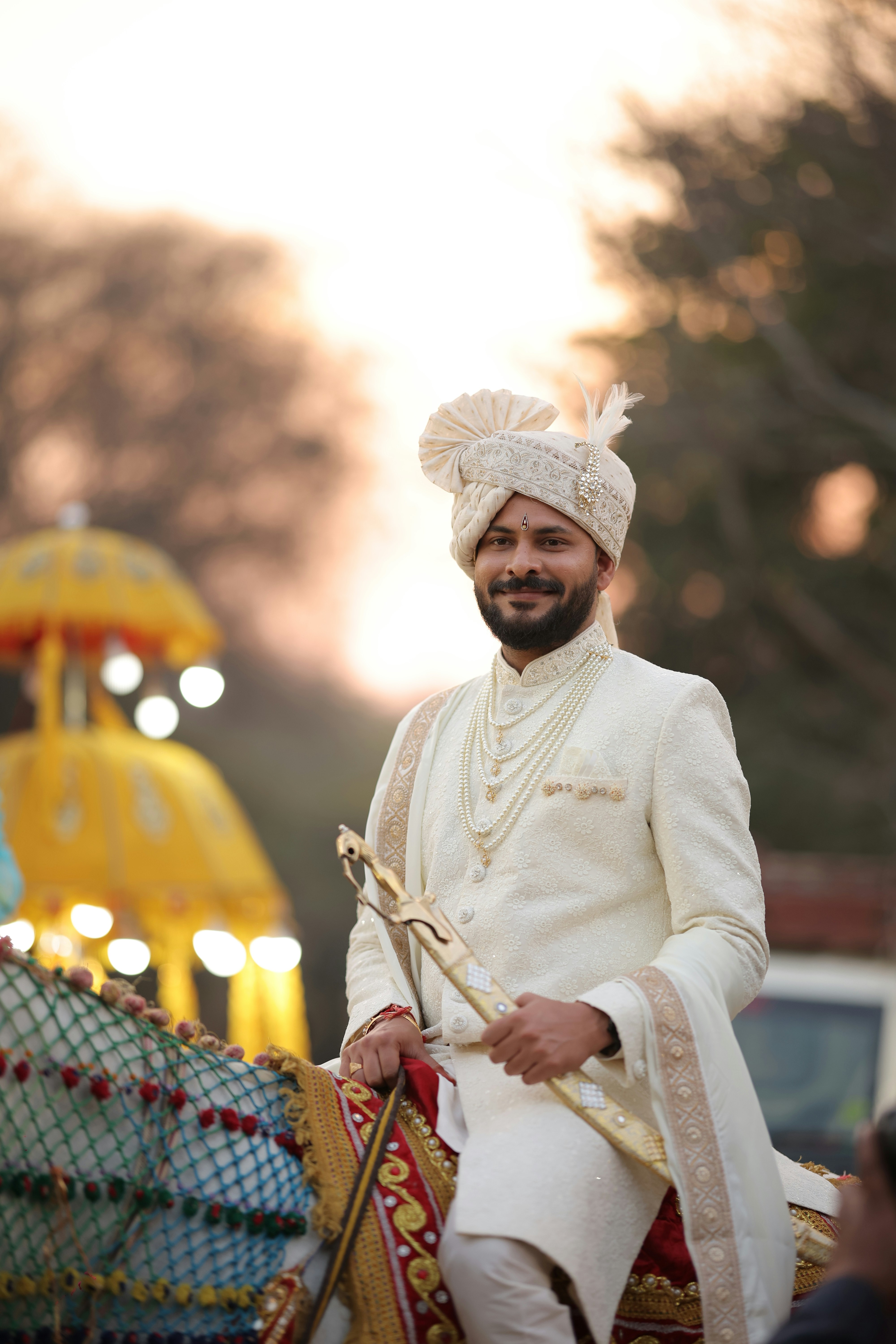 The groom, riding a white horse decorated with vibrant and colorful ornaments, raises his hand in a victorious gesture, his face beaming with joy and anticipation as the baraat makes its way to the wedding venue. | A man in a turban riding a horse