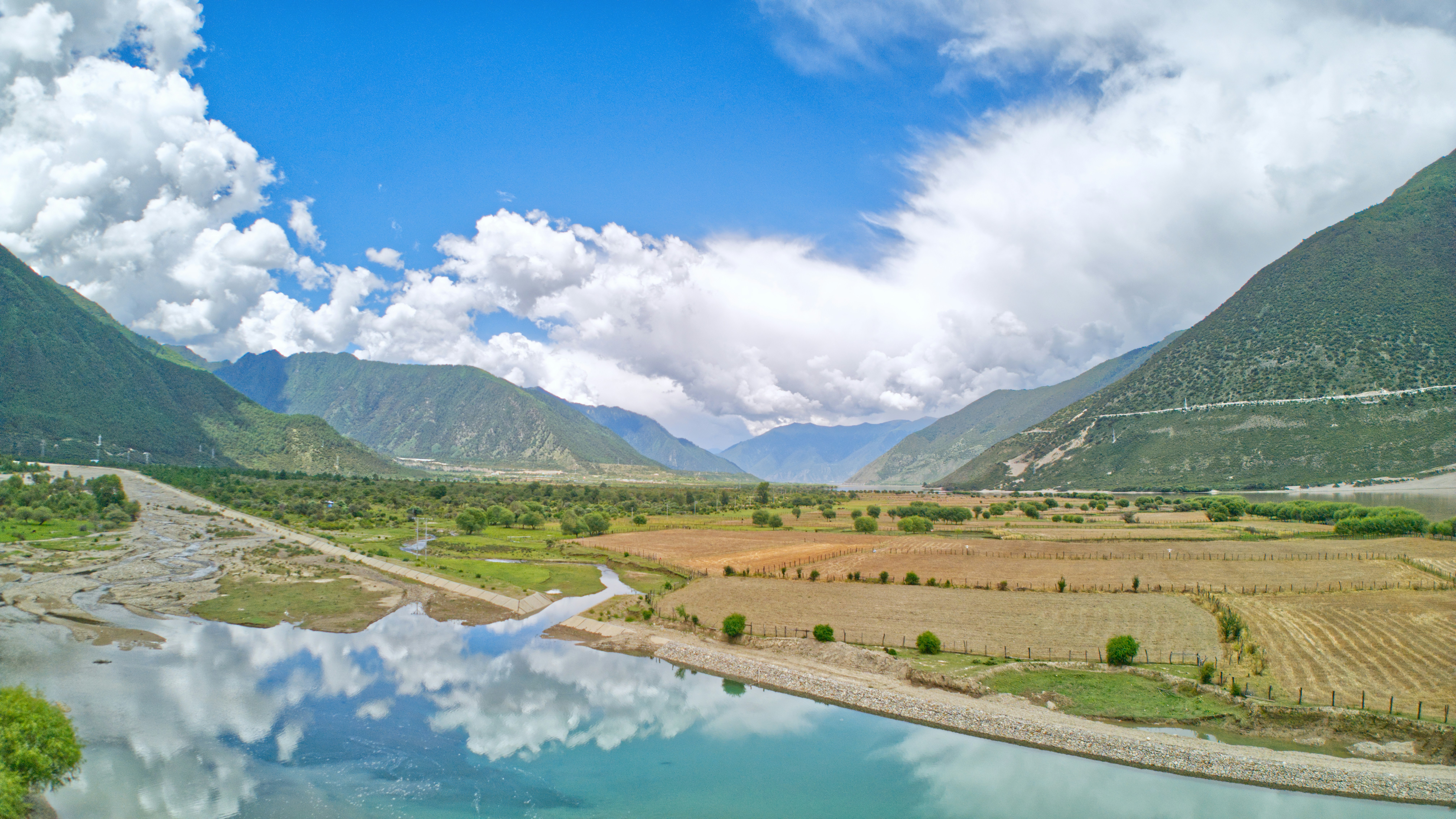 River reflecting clouds and mountains in a lush valley under a vibrant blue sky.