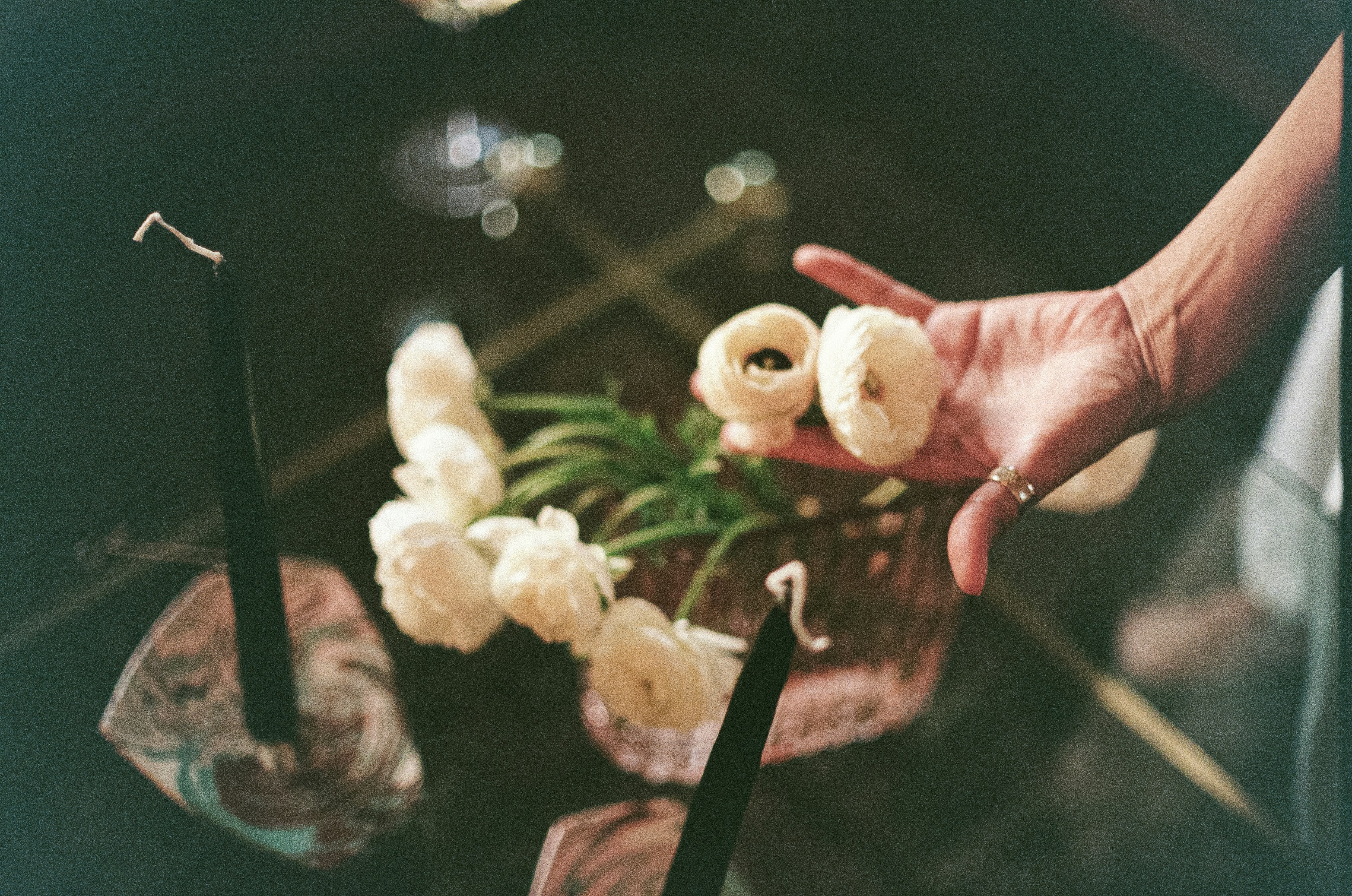 Warm-toned top-down photograph of two dark candle holders and a white floral arrangement; a hand enters from the right, adding a human element to the composition.
