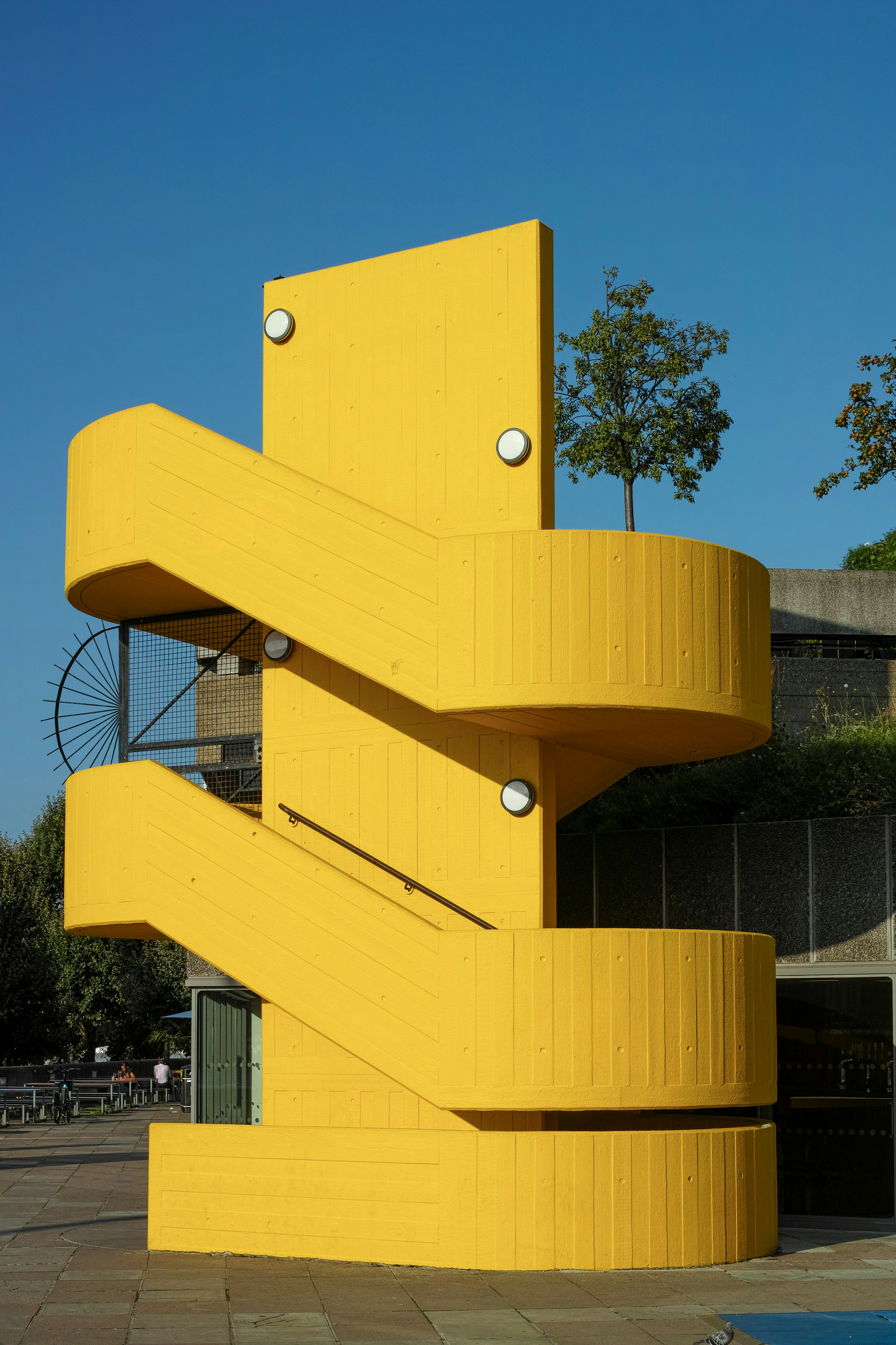 Photograph of a bright yellow spiraling staircase sculpture outdoors against a clear blue sky. The structure sits near modern architecture with a tree peeking from the top.